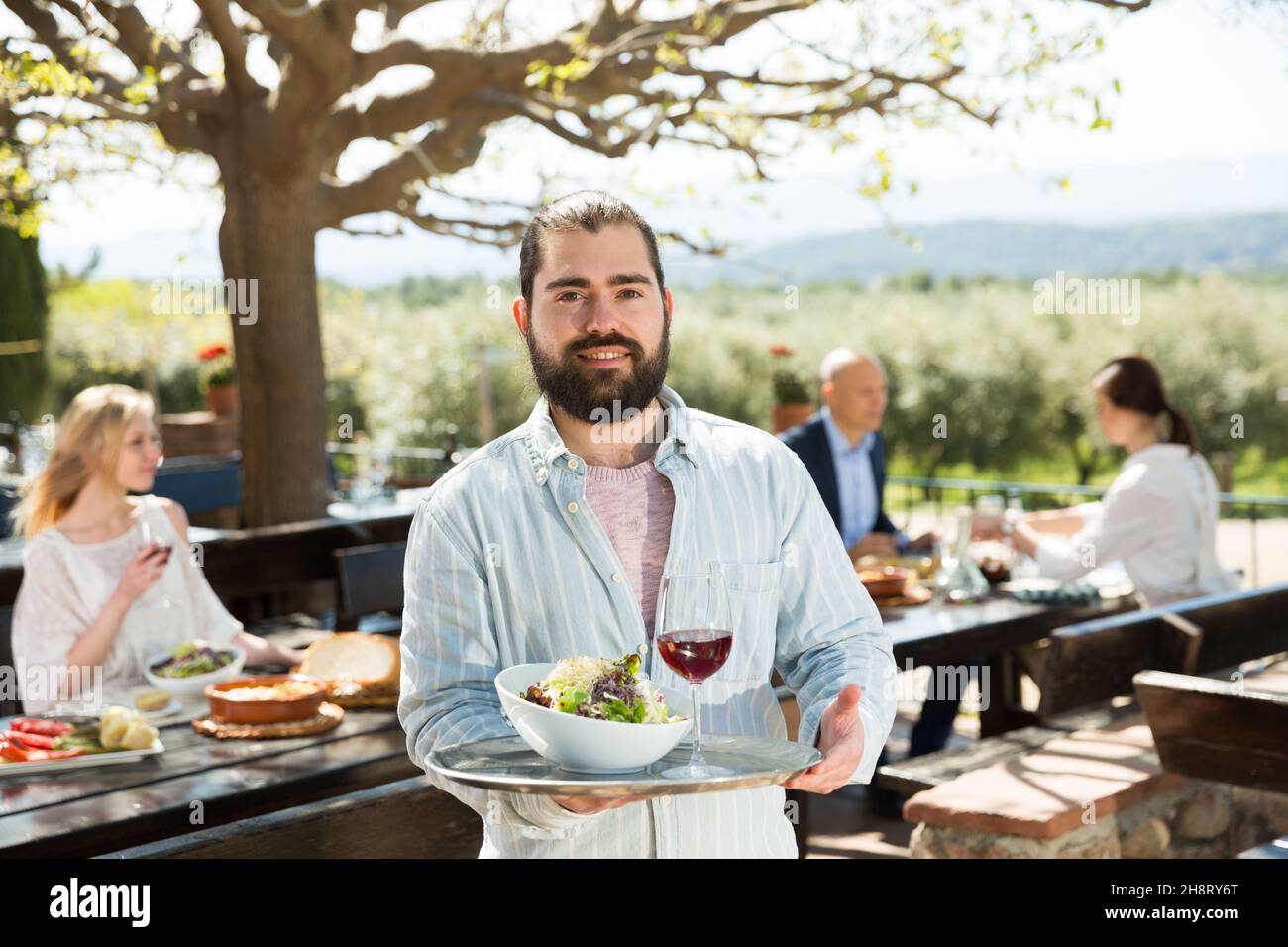 Positive male waiter welcoming guests to country outdoor restaurant ...