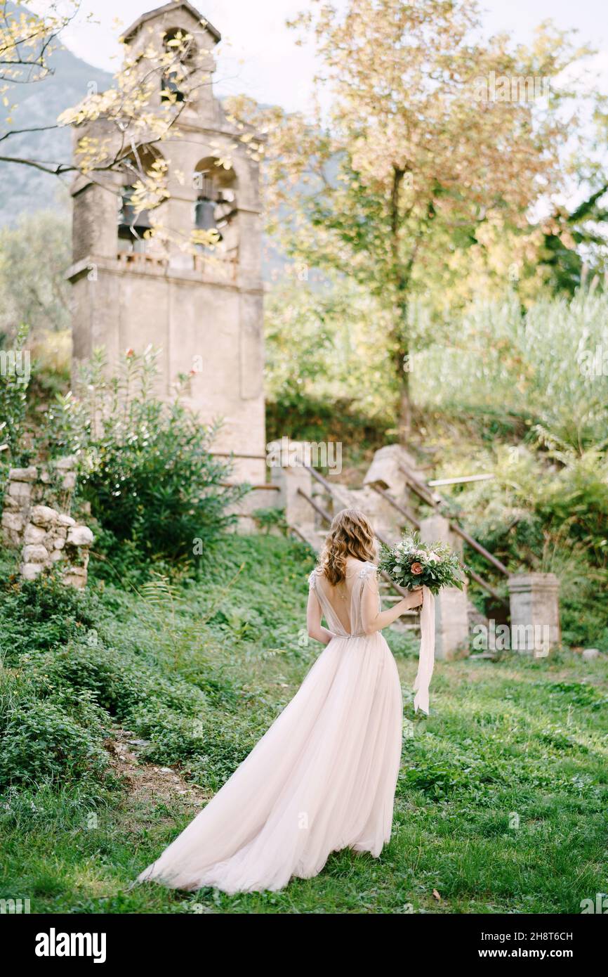 Bride with a bouquet walks along the green grass to the ancient chapel ...