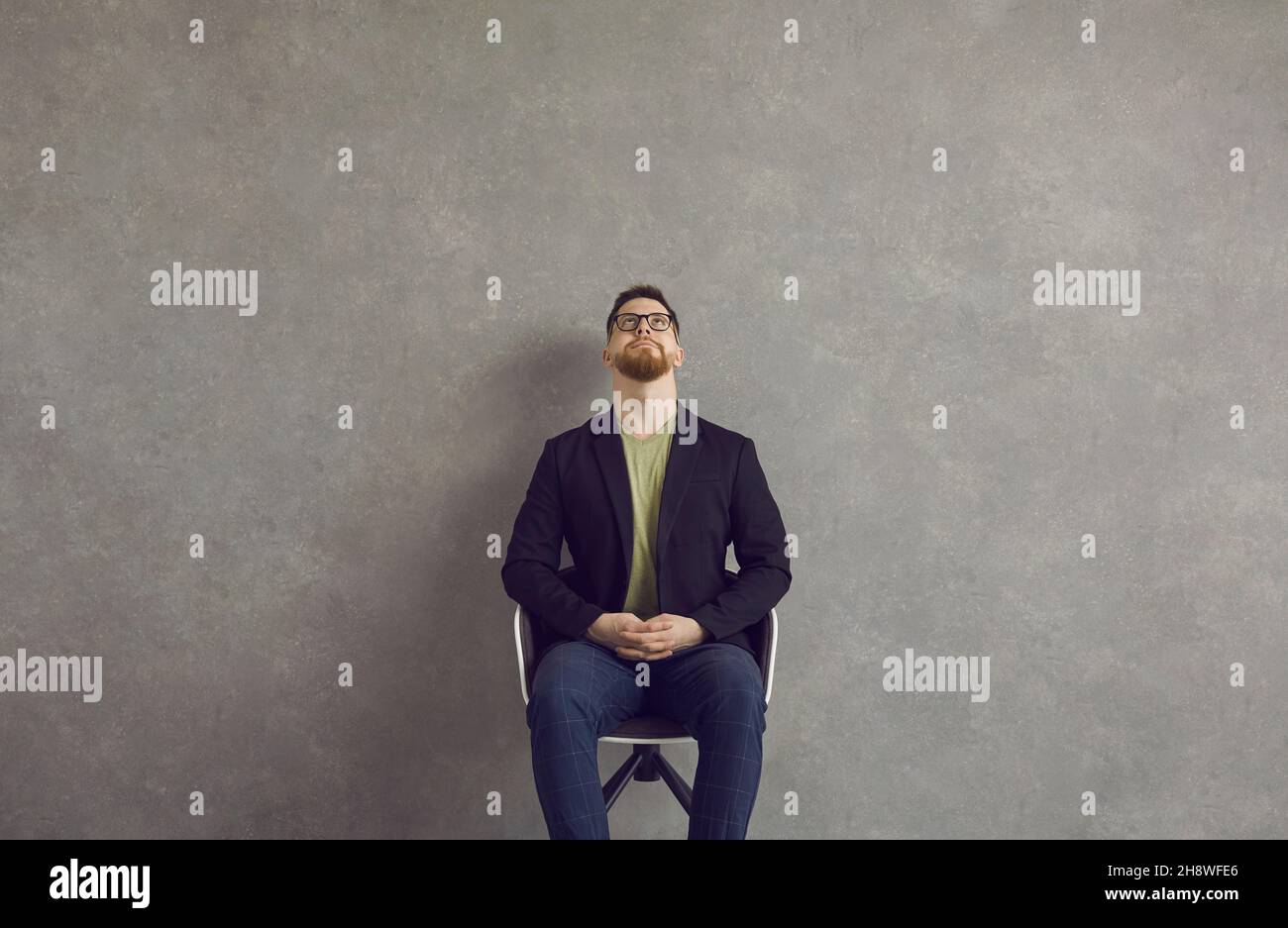 Young man sitting in chair near wall, looking up, thinking or imagining ...