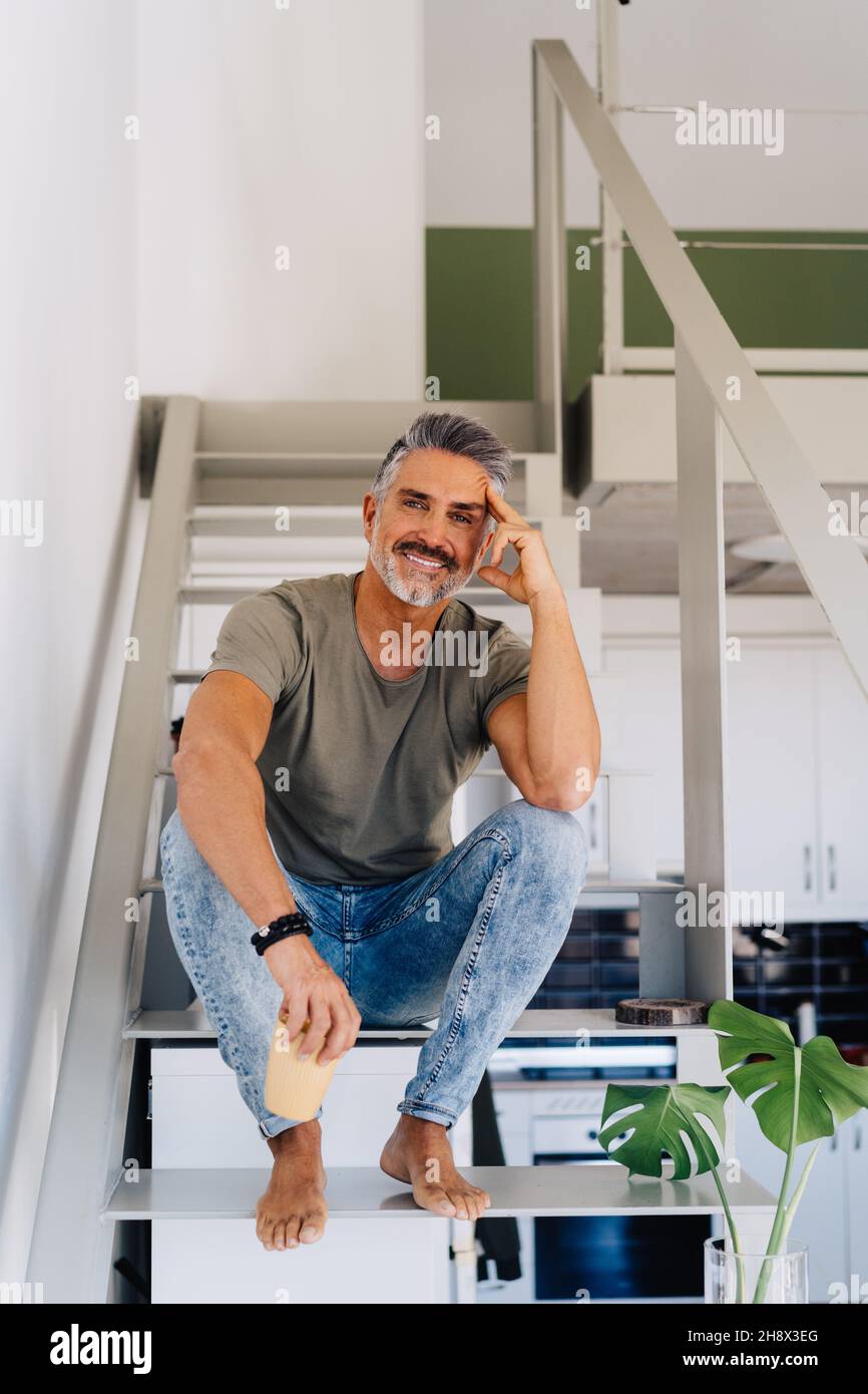 Elderly cheerful barefoot man sitting on ladder and looking at camera ...
