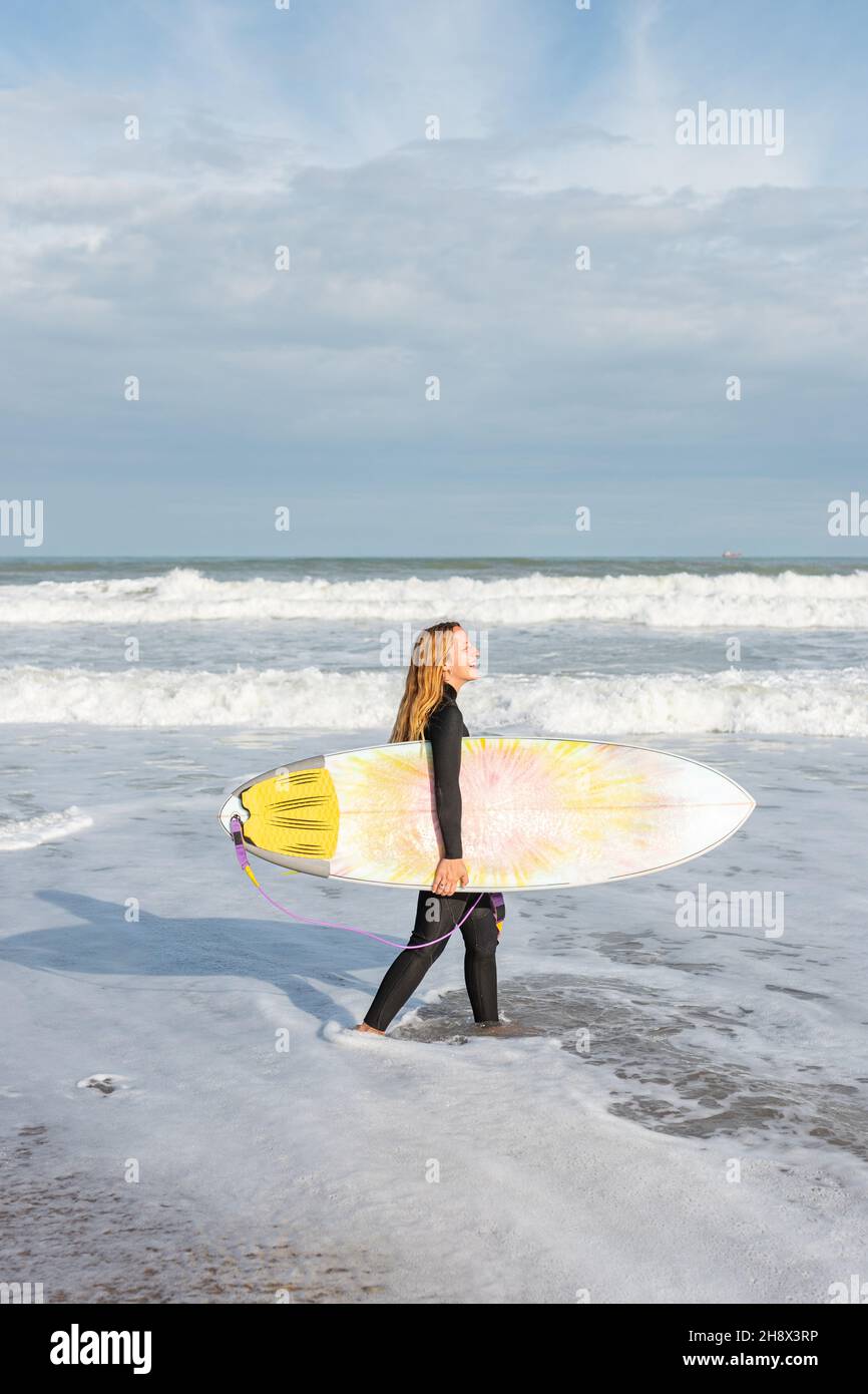 Full body side view of sportive female surfer in black wetsuit walking ...