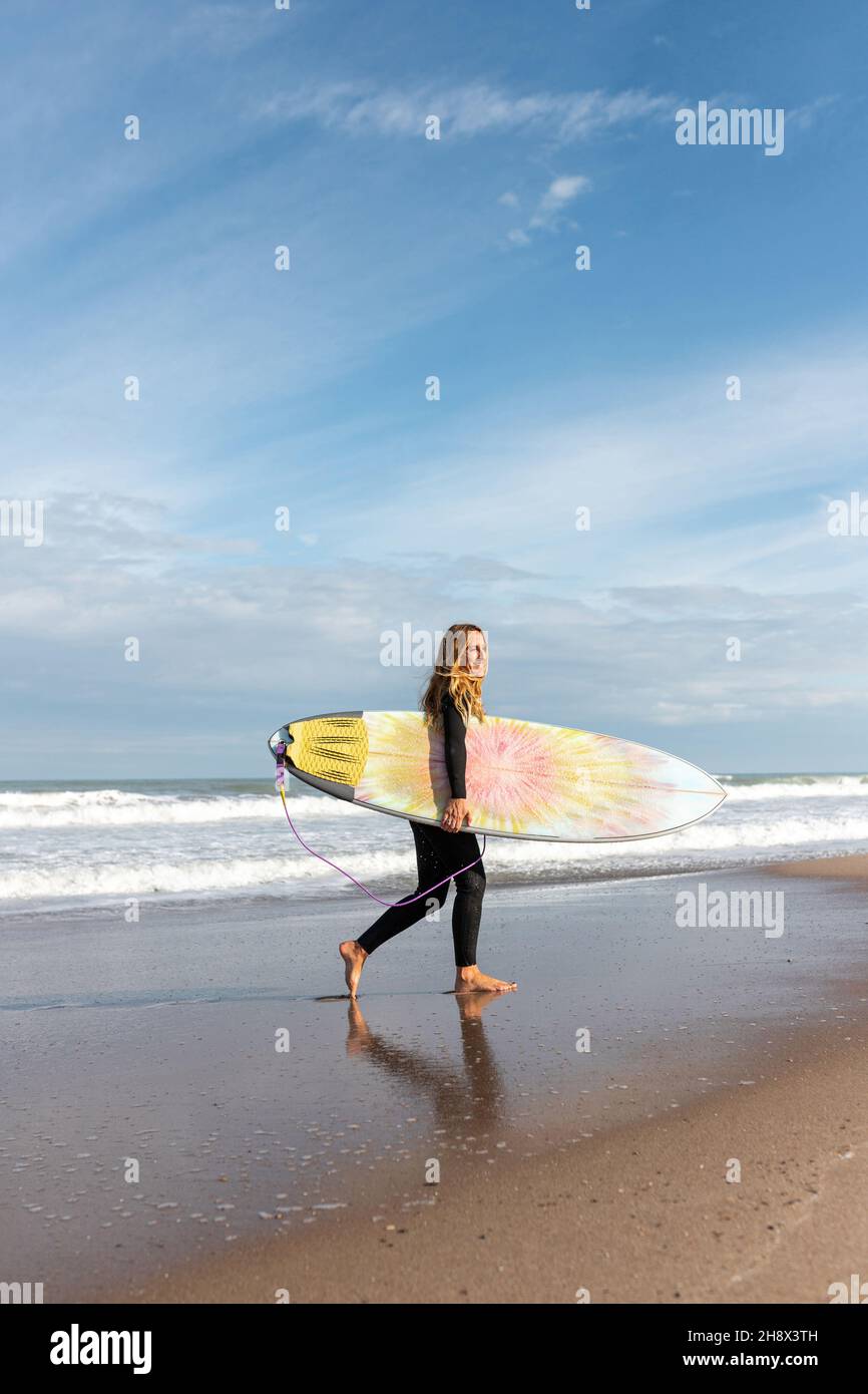 Full body side view of sportive female surfer in black wetsuit walking ...