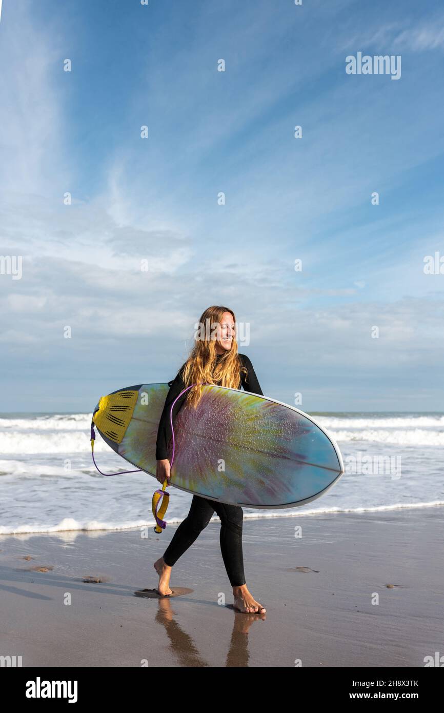 Full body side view of sportive female surfer in black wetsuit walking ...