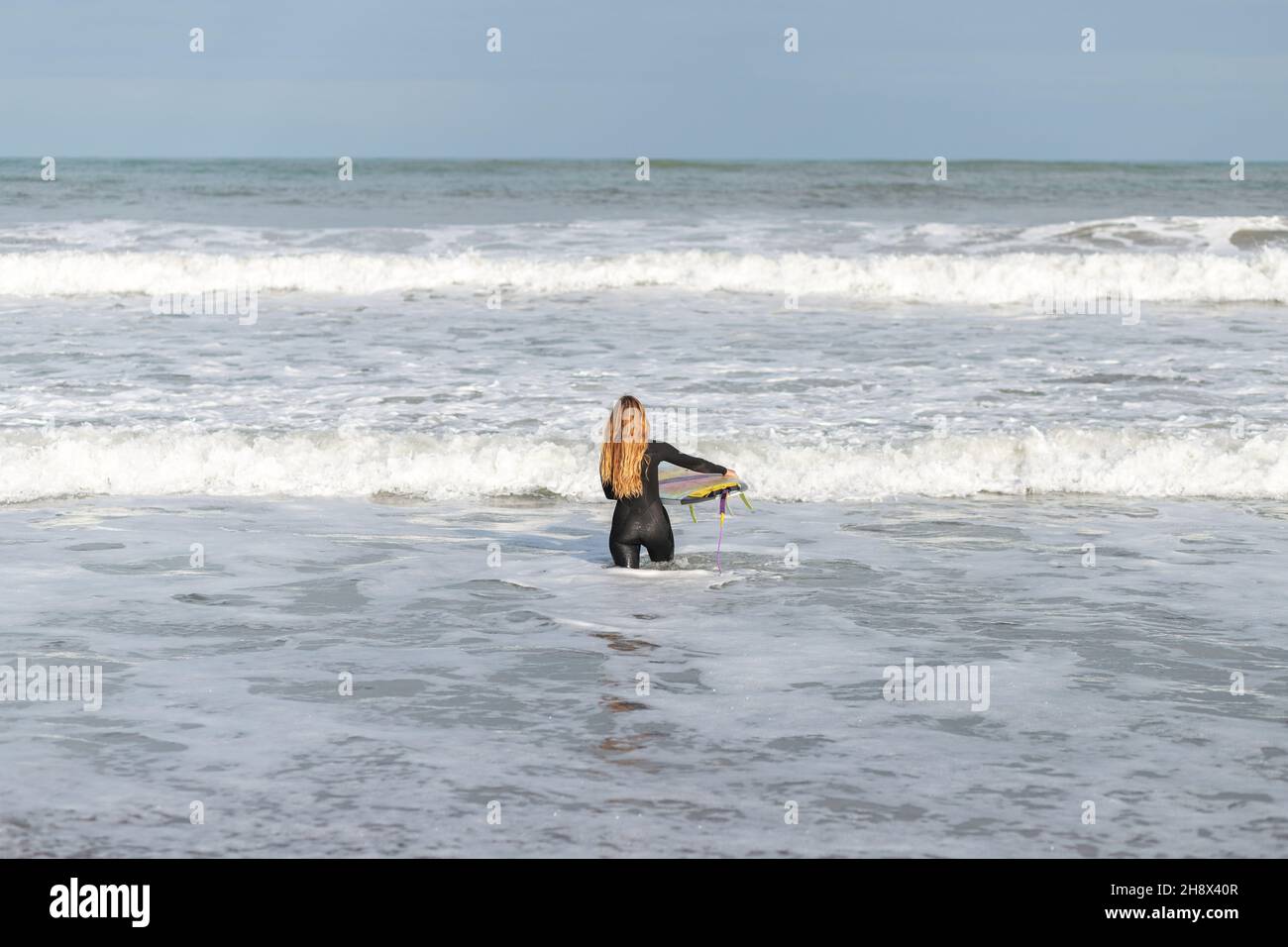Back view of anonymous Woman surfer in wetsuit with surfboard in waving ...