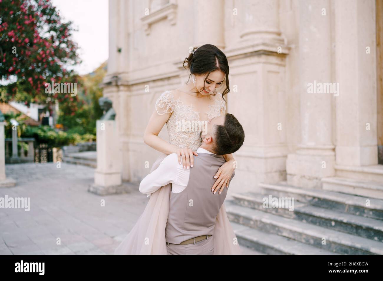 Groom holds bride in his arms in front of the marble facade of the old ...