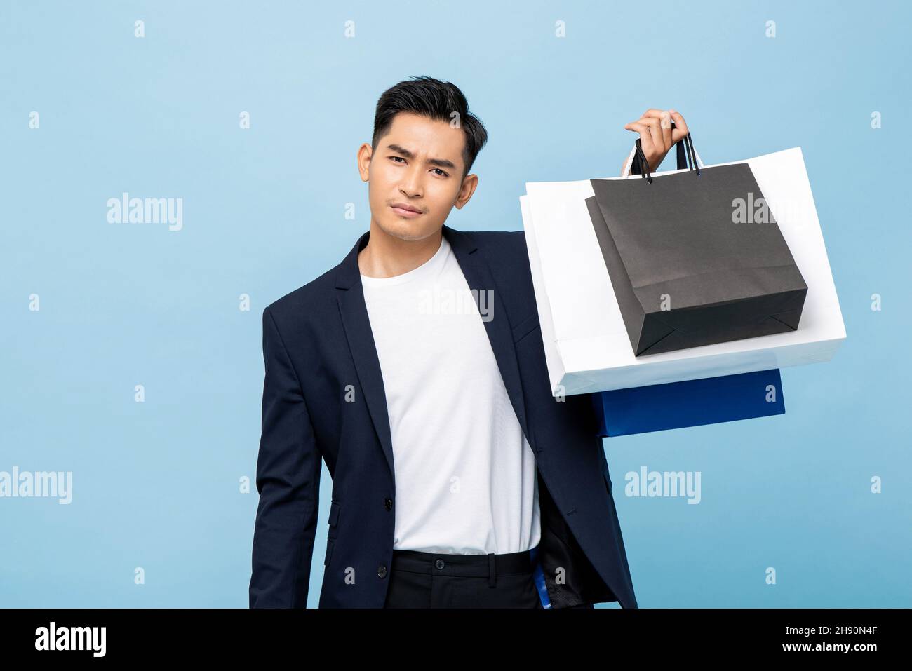 Young doubtful Asian man holding shopping bags in light blue isolated ...