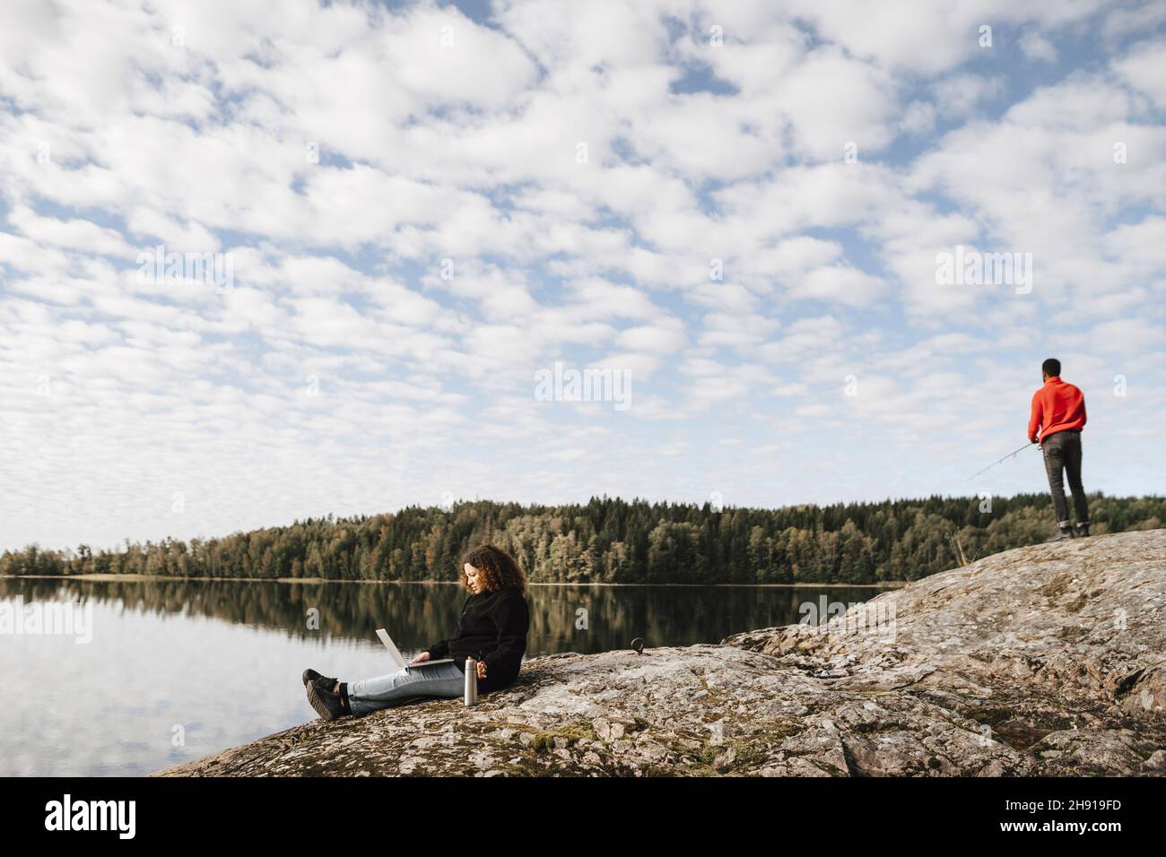 Woman working remotely on laptop while boyfriend fishing at lakeshore ...
