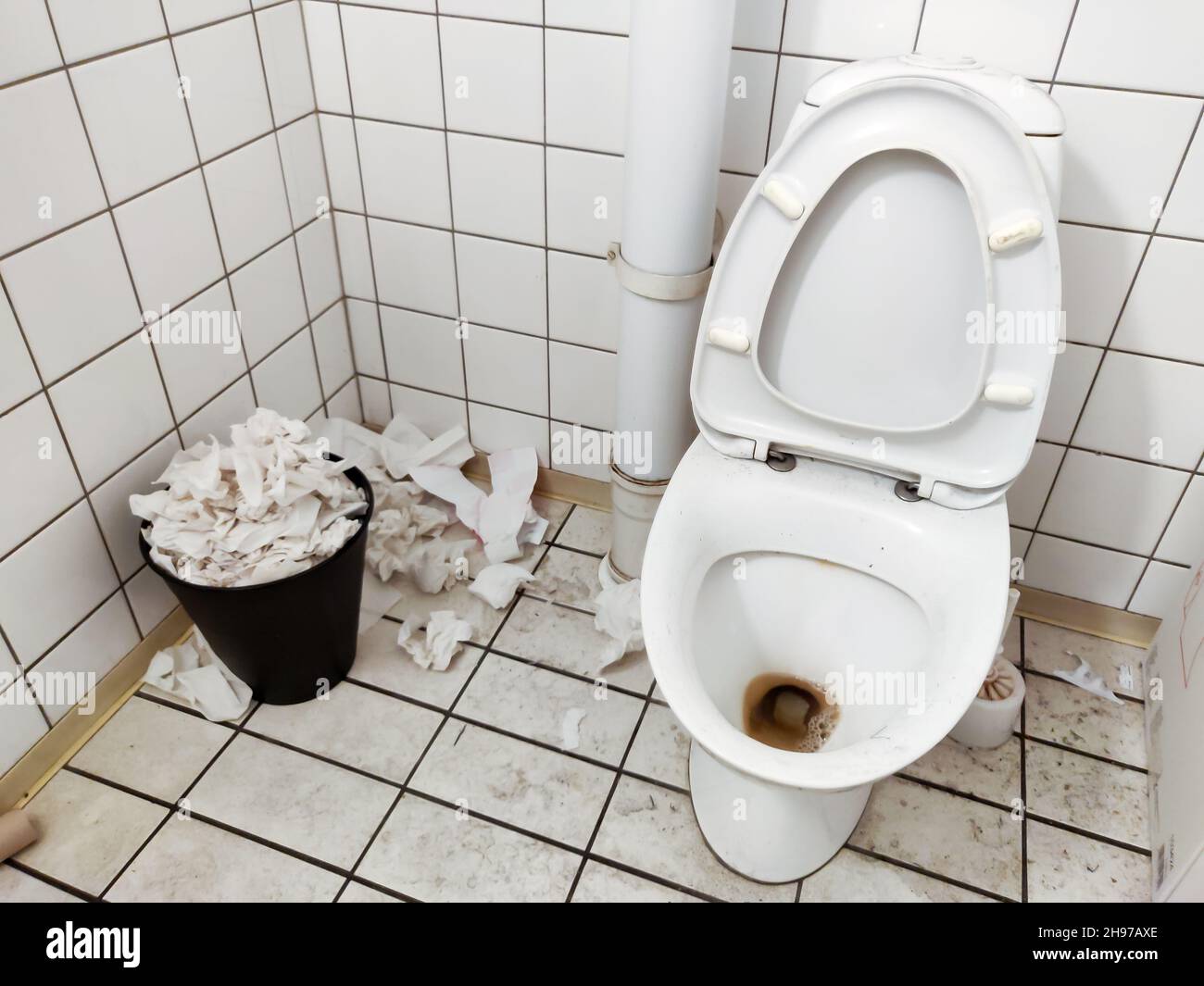 A white uncleaned toilet with white tiled floor and a bin full of ...