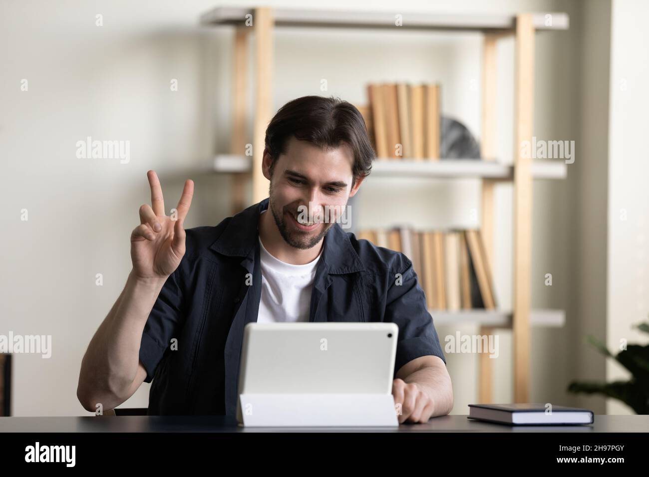 Positive millennial employee guy, tablet user waving hello at screen ...