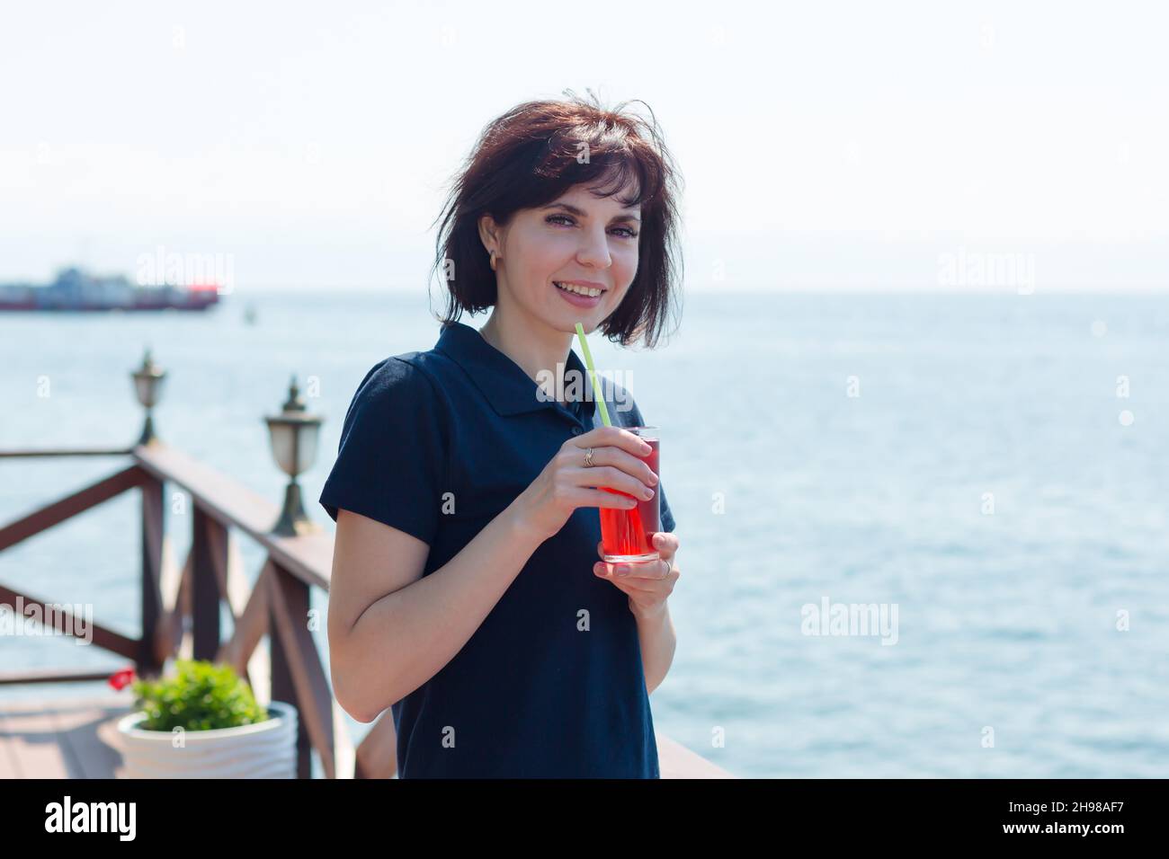 Smiling brunette in a blue dress on the pier with a glass of strawberry juice Stock Photo - Alamy