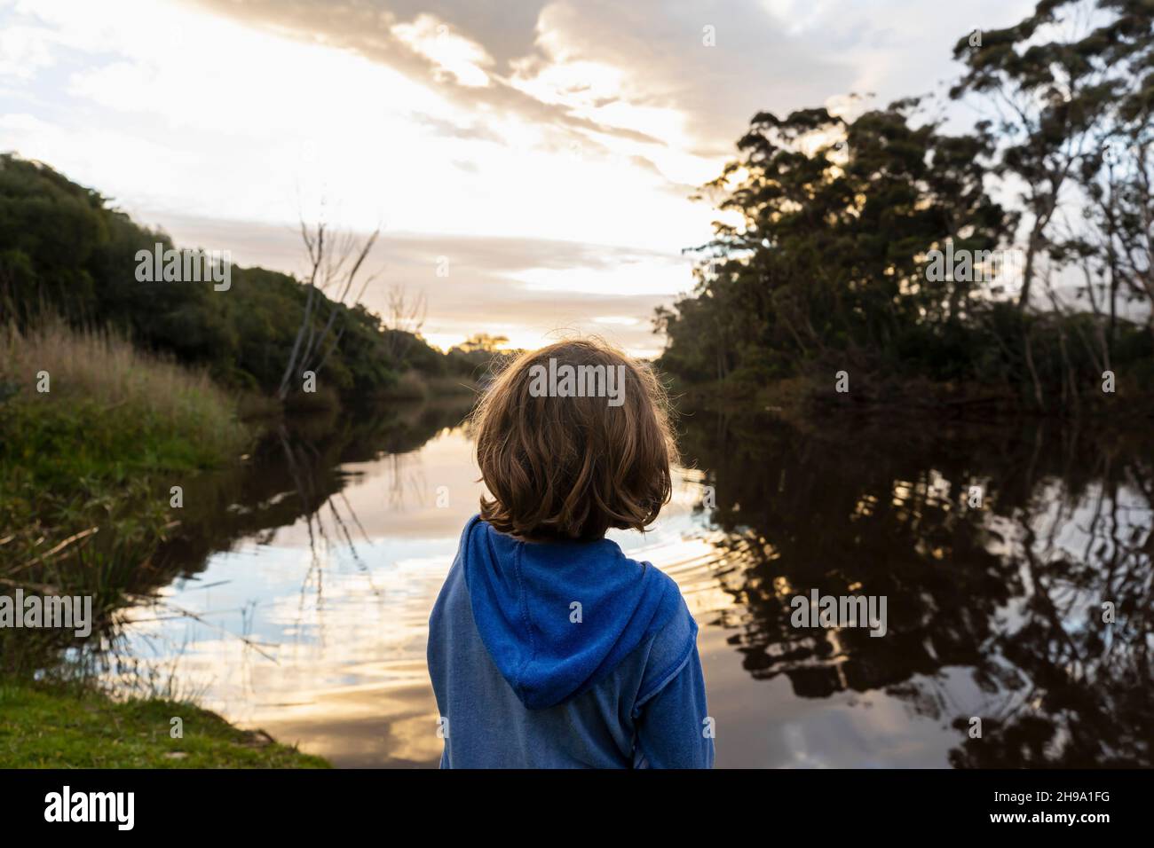 A young boy standing by a river at dusk, sky reflections in the flat ...
