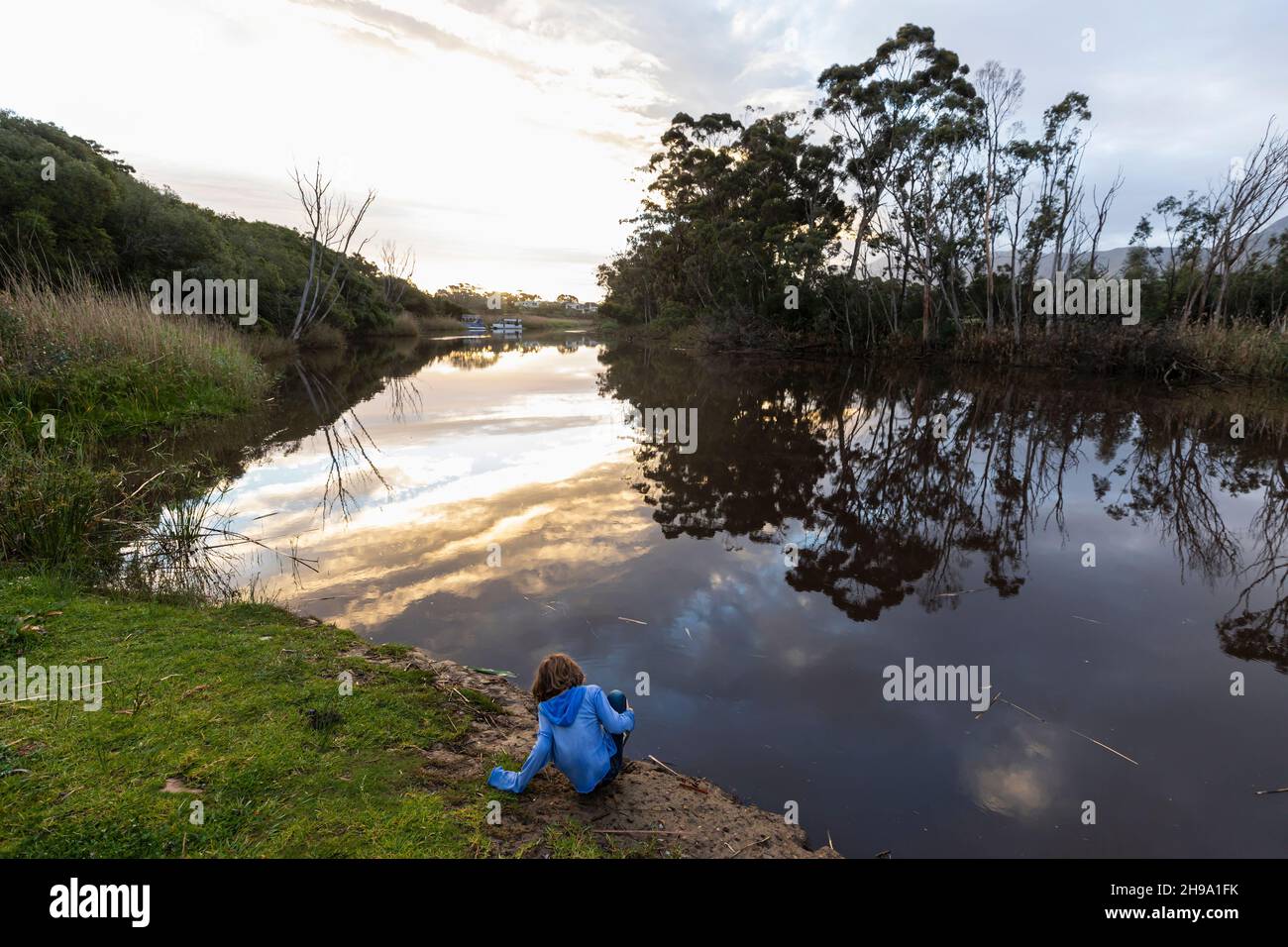 A young boy standing by a river at dusk, sky reflections in the flat ...