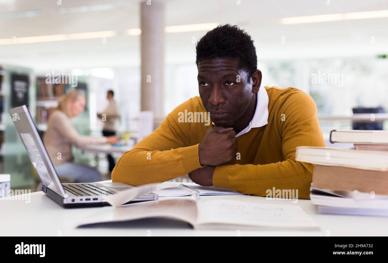 Thoughtful african-american man working with textbook and laptop in ...