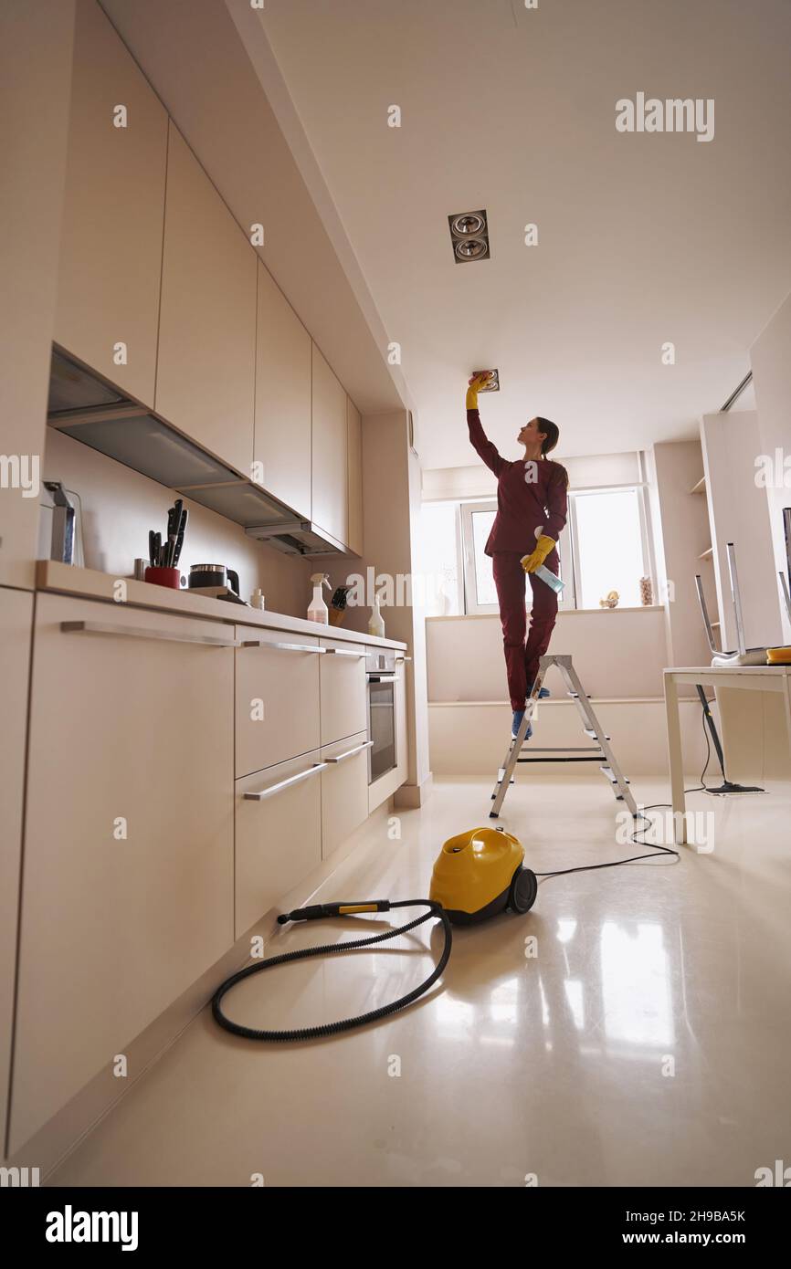 Female cleaner in rubber gloves standing on step-ladder and dusting ...
