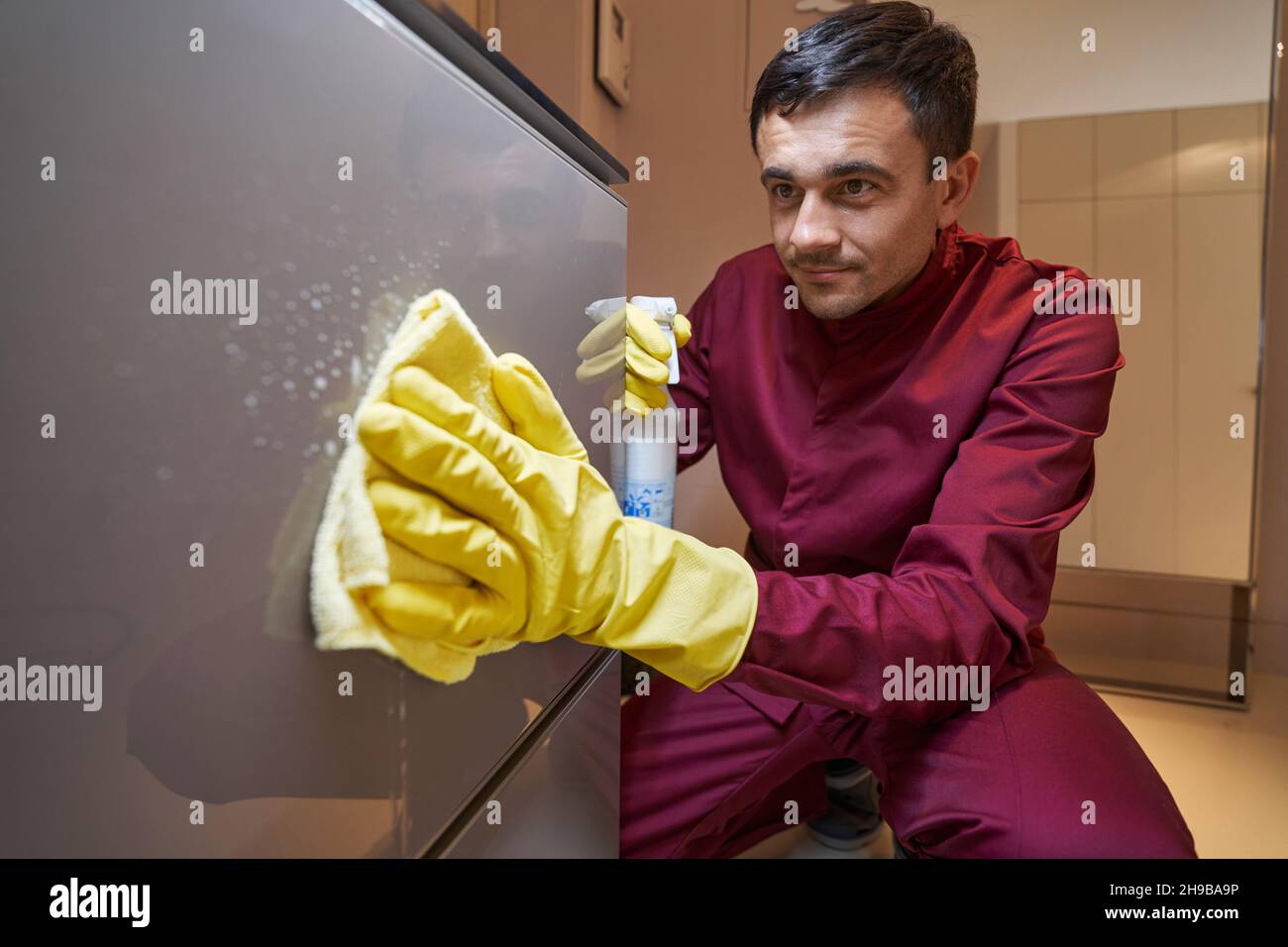 Male janitor in rubber gloves sitting on haunches and cleaning piece of ...