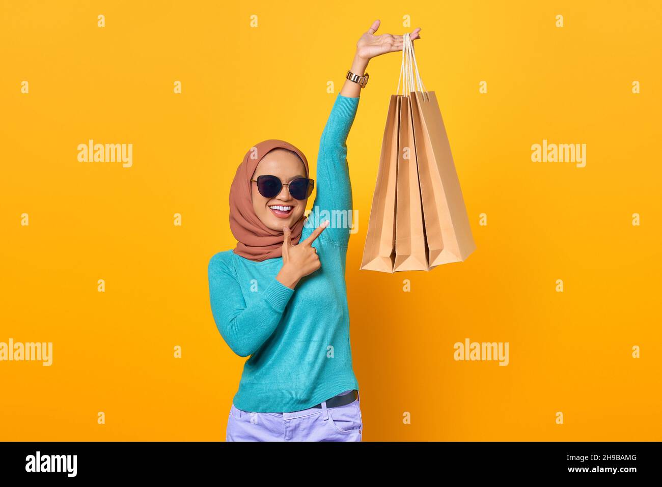 Smiling young Asian woman pointing fingers at shopping bags on yellow ...