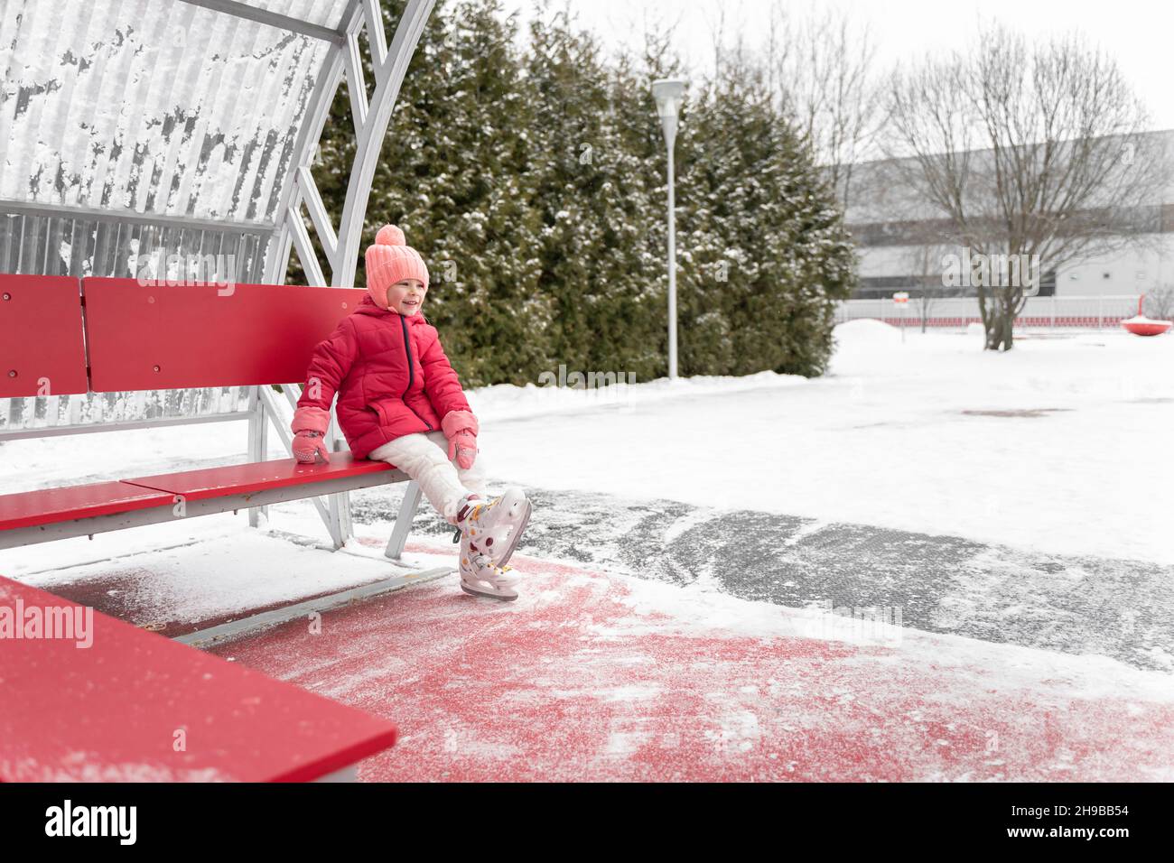 adorable kid girl with skates sits on a bench near the winter outdoor ...