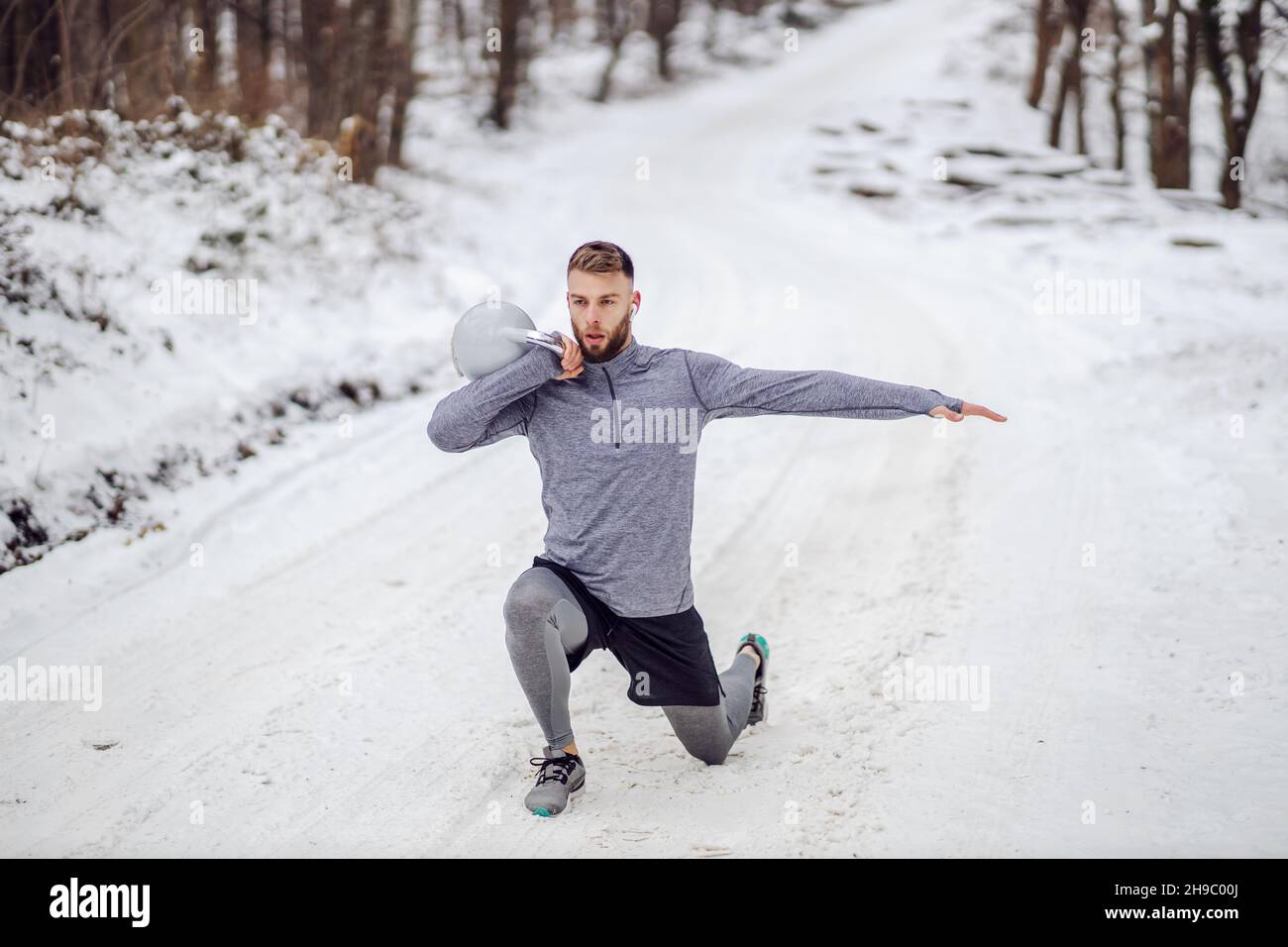 Fit sportsman lifting kettlebell while doing lunges on snowy path in ...