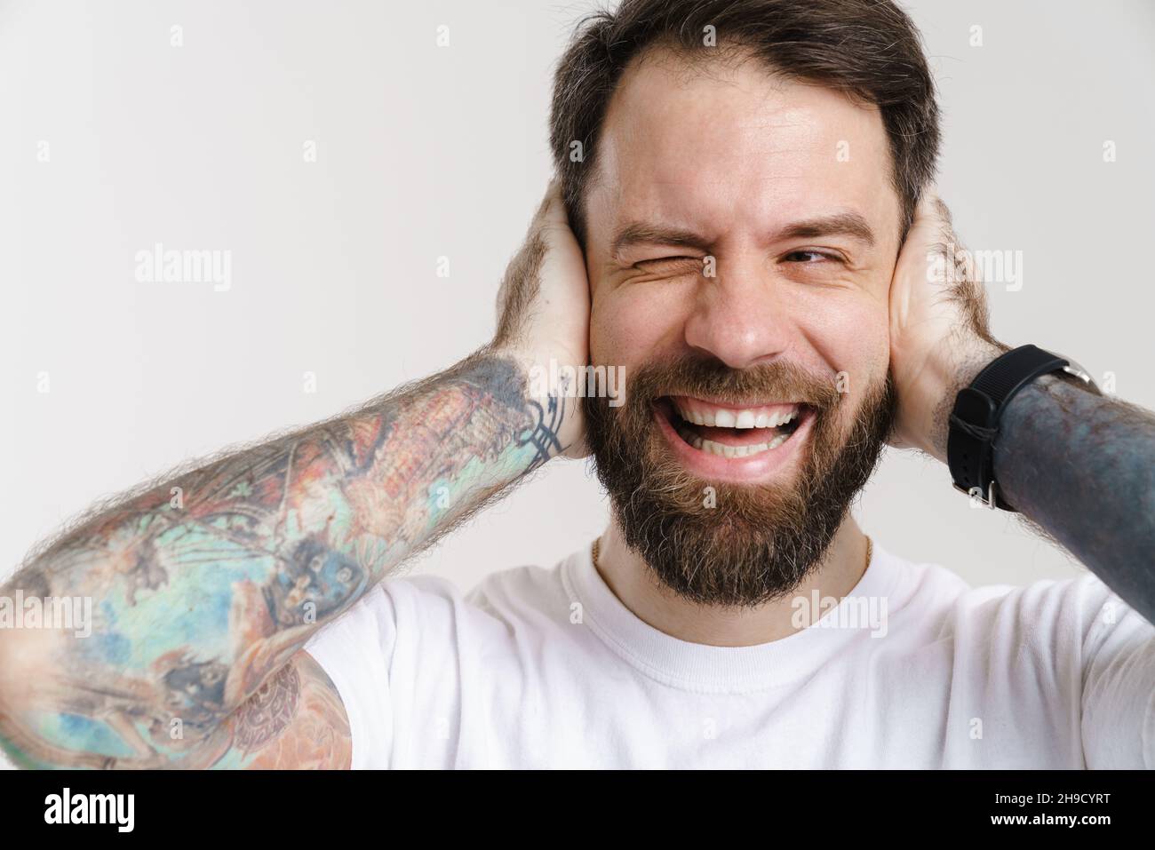Bearded white man with tattoo laughing and covering his ears isolated ...