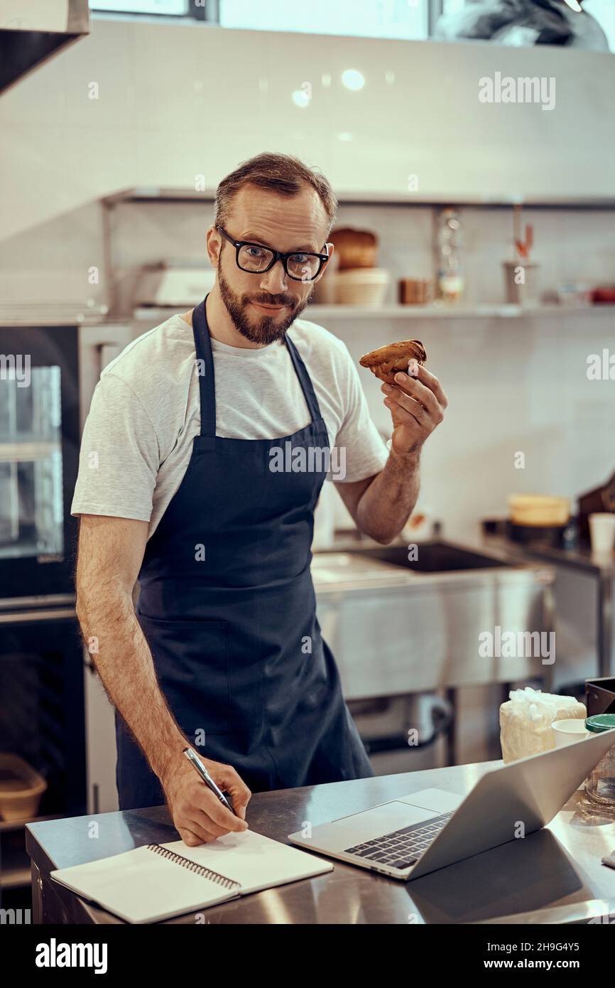 Male cafe owner holding pastry and writing in notebook Stock Photo - Alamy