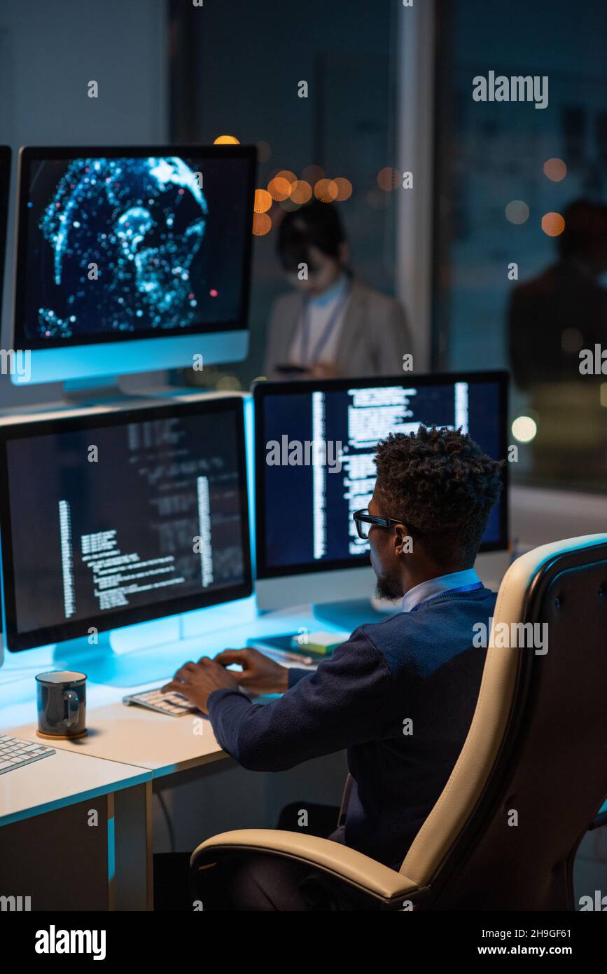 Young African developer sitting in armchair by desk and typing while looking at coded data on ...