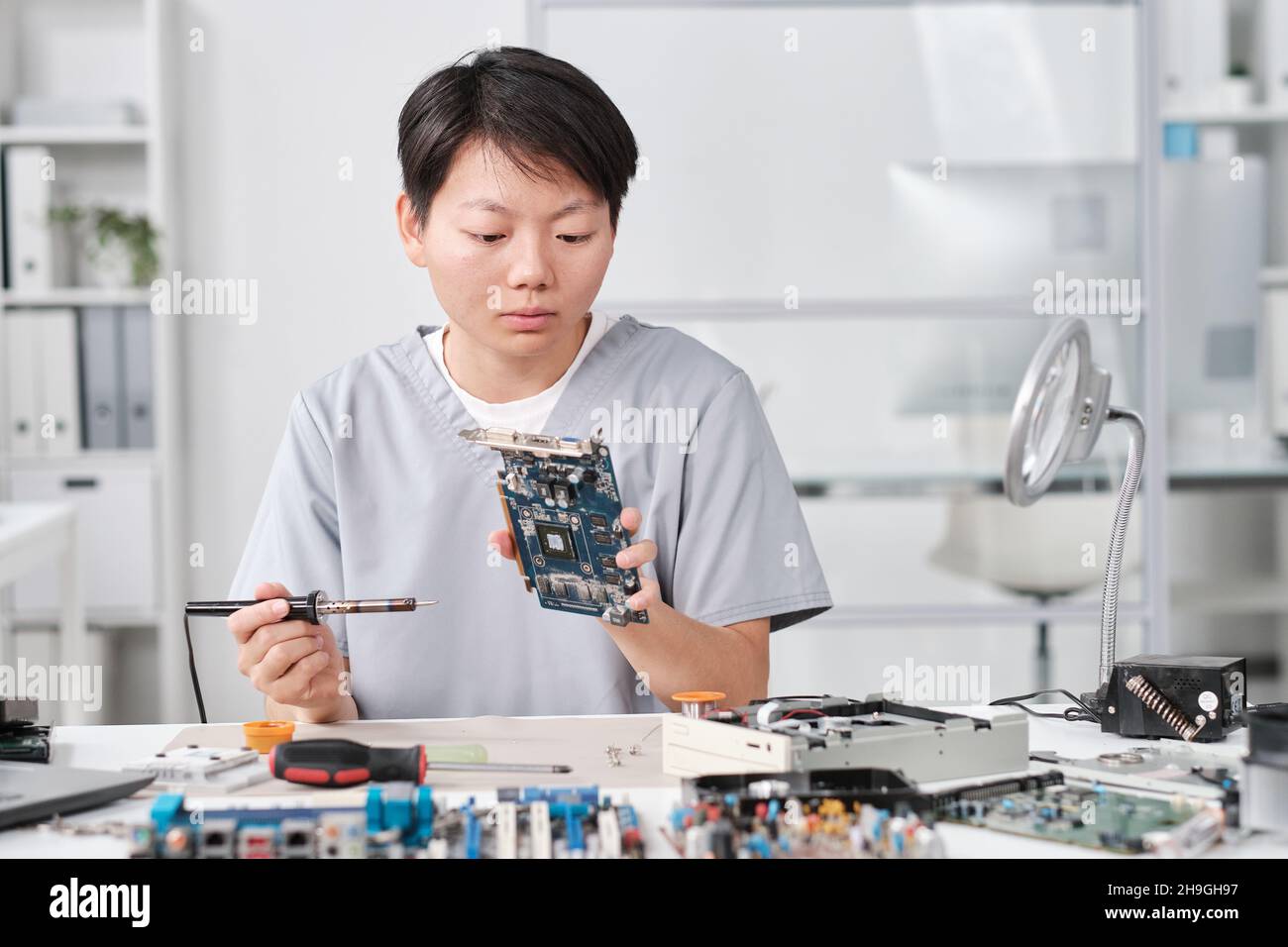 Young serious Asian woman with soldering iron looking at hardware while ...