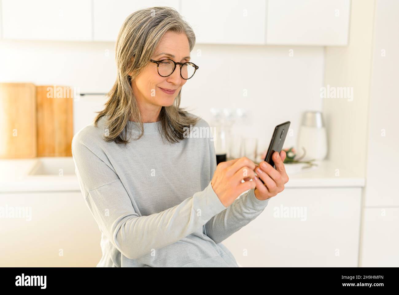 Happy smiling mature woman with gray hair holds smartphone sitting in the kitchen. Charming lady ...