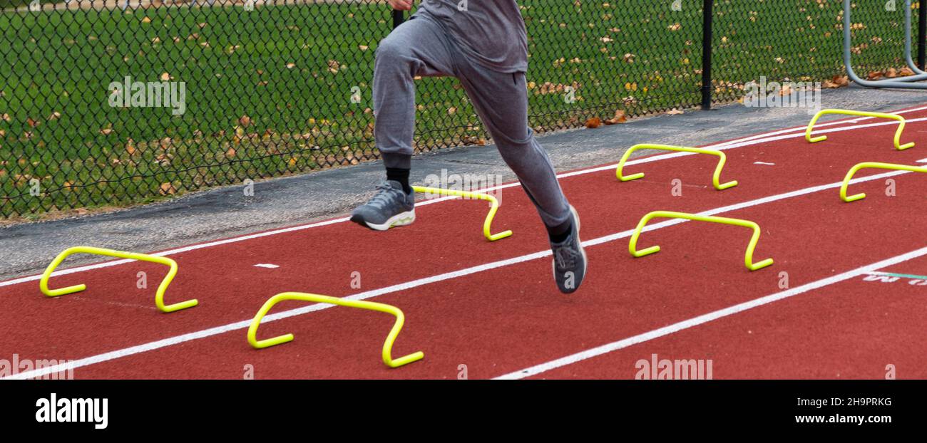A high school runner is learning speed acceleration by running over yellow mini hurdles on a red ...