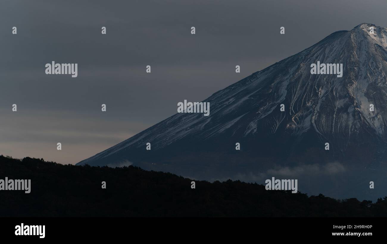 Shot of a snowy peak of Mount Fuji after sunset under the cloudy sky ...