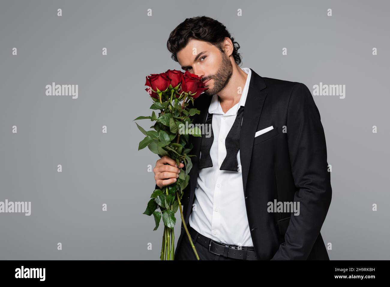 bearded man in suit smelling red roses while looking at camera isolated ...