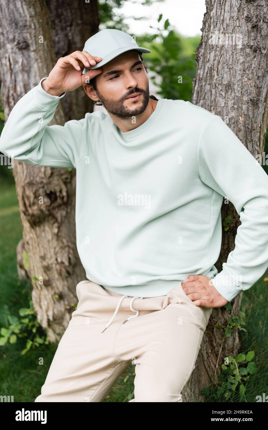 bearded man adjusting cap and standing near tree trunks in park Stock ...