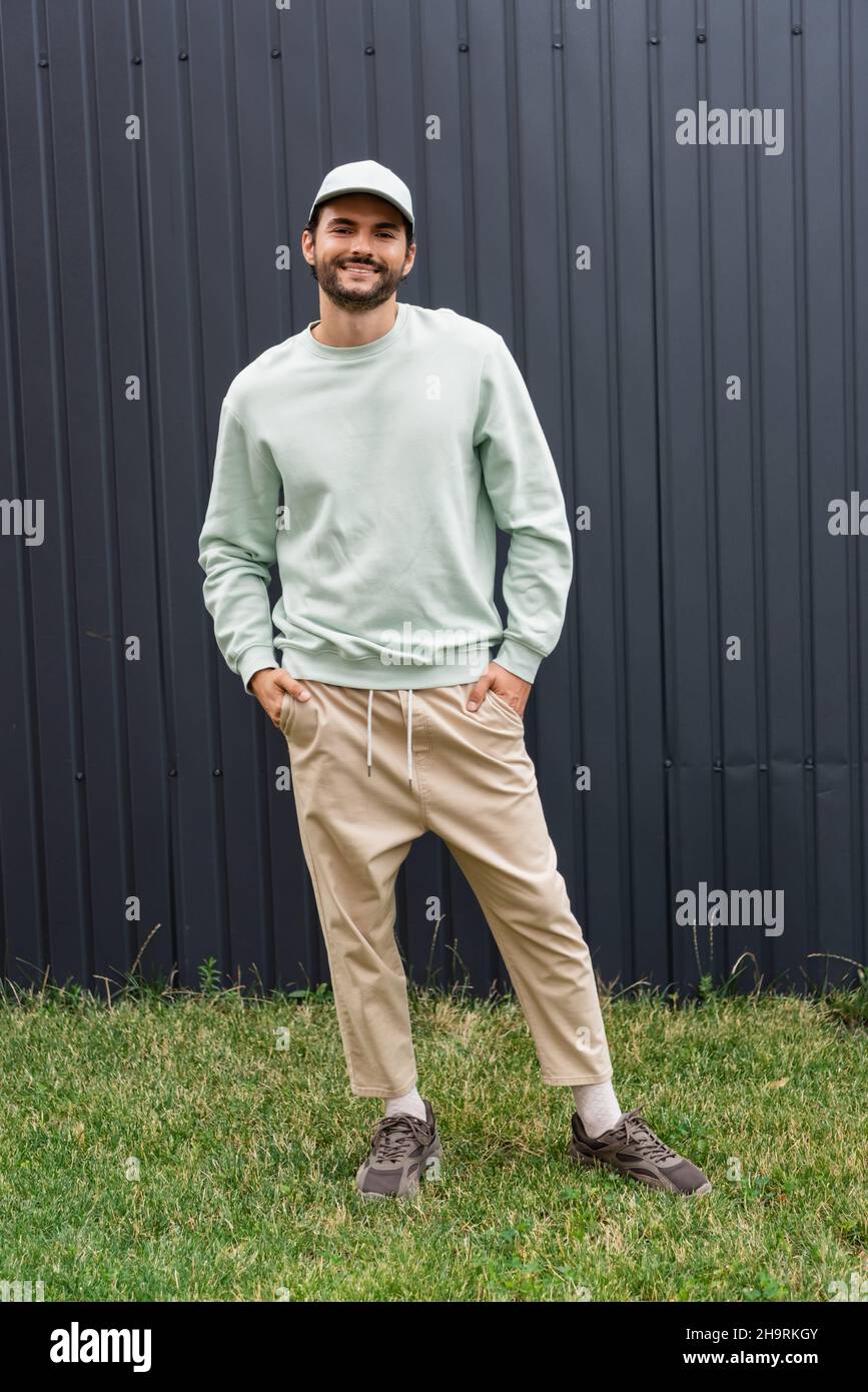 full length of smiling man in baseball cap posing with hands in pockets ...