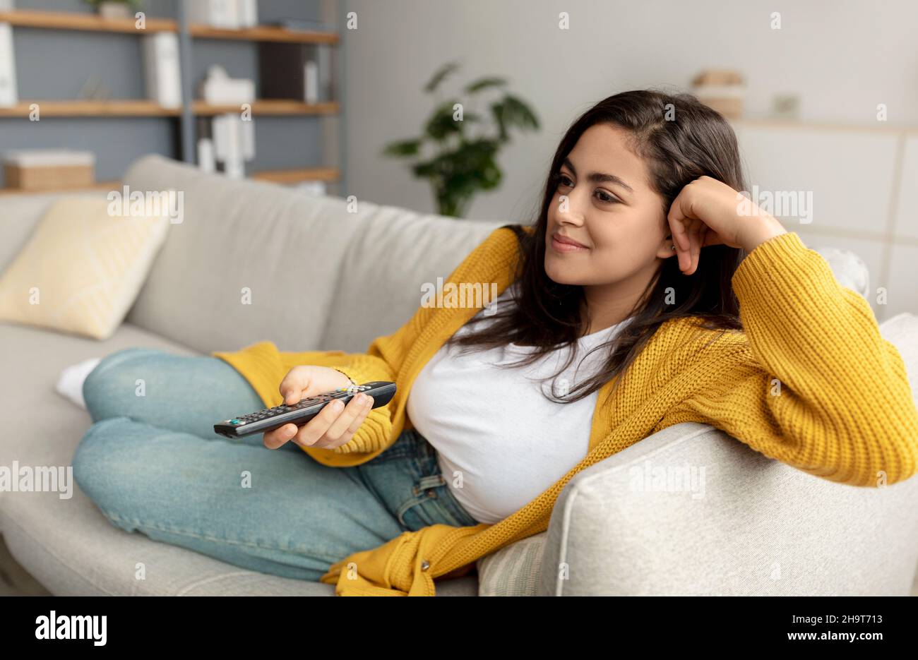 Pretty arab lady with remote control watching TV on couch at home ...