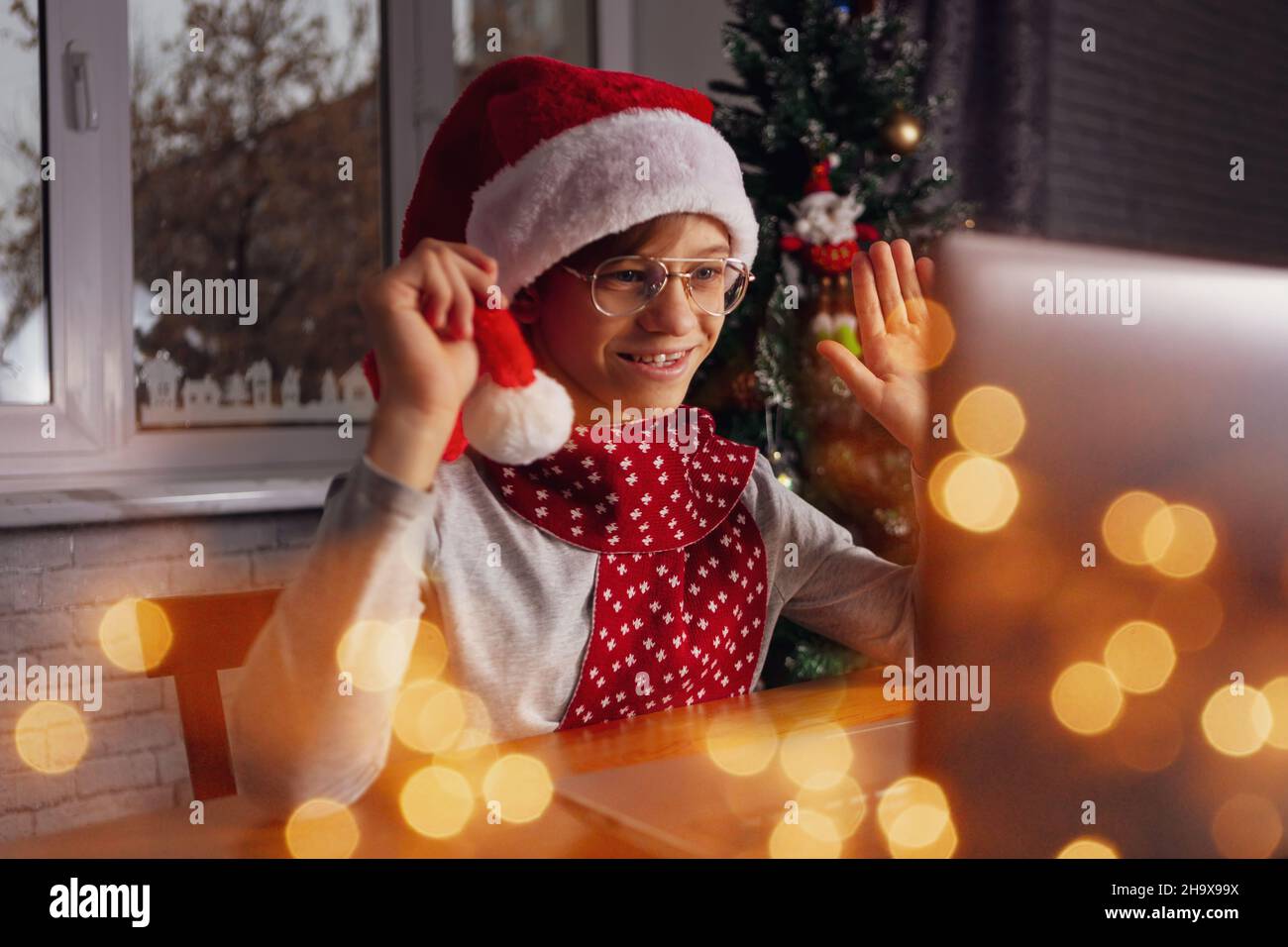 A smiling boy sitting in front of laptop screen wearing red Santa Claus ...