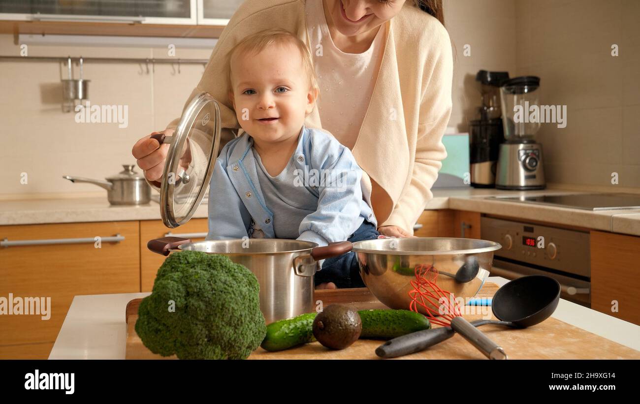 Happy smiling mother showing pans and kitchen utensils to her baby son ...