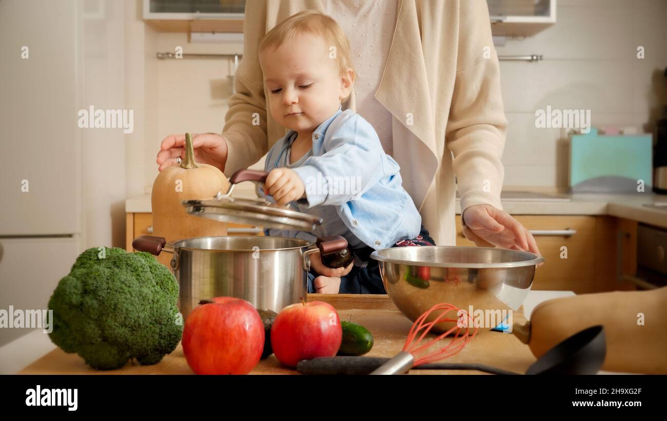Little baby boy sitting on kitchen table and looking inside cooking pan ...