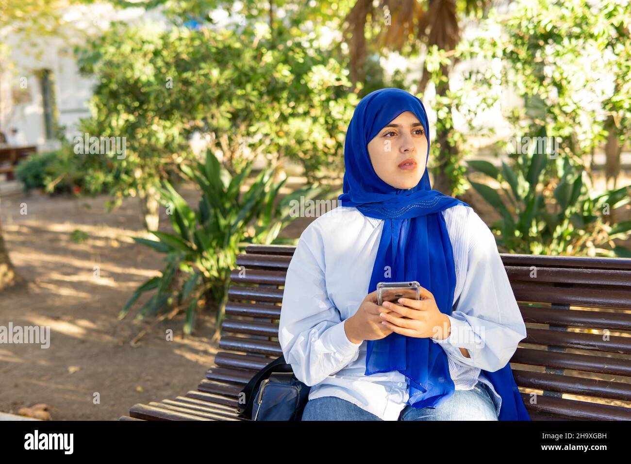 Young Muslim woman wearing hijab sitting on park bench using the phone ...