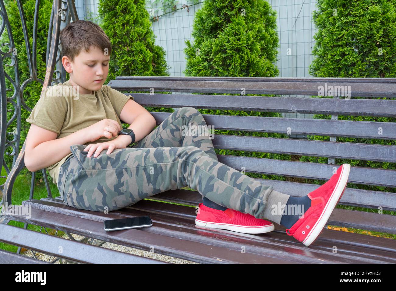 A teenage boy sitting on a bench looking at his watch. A boy in the ...