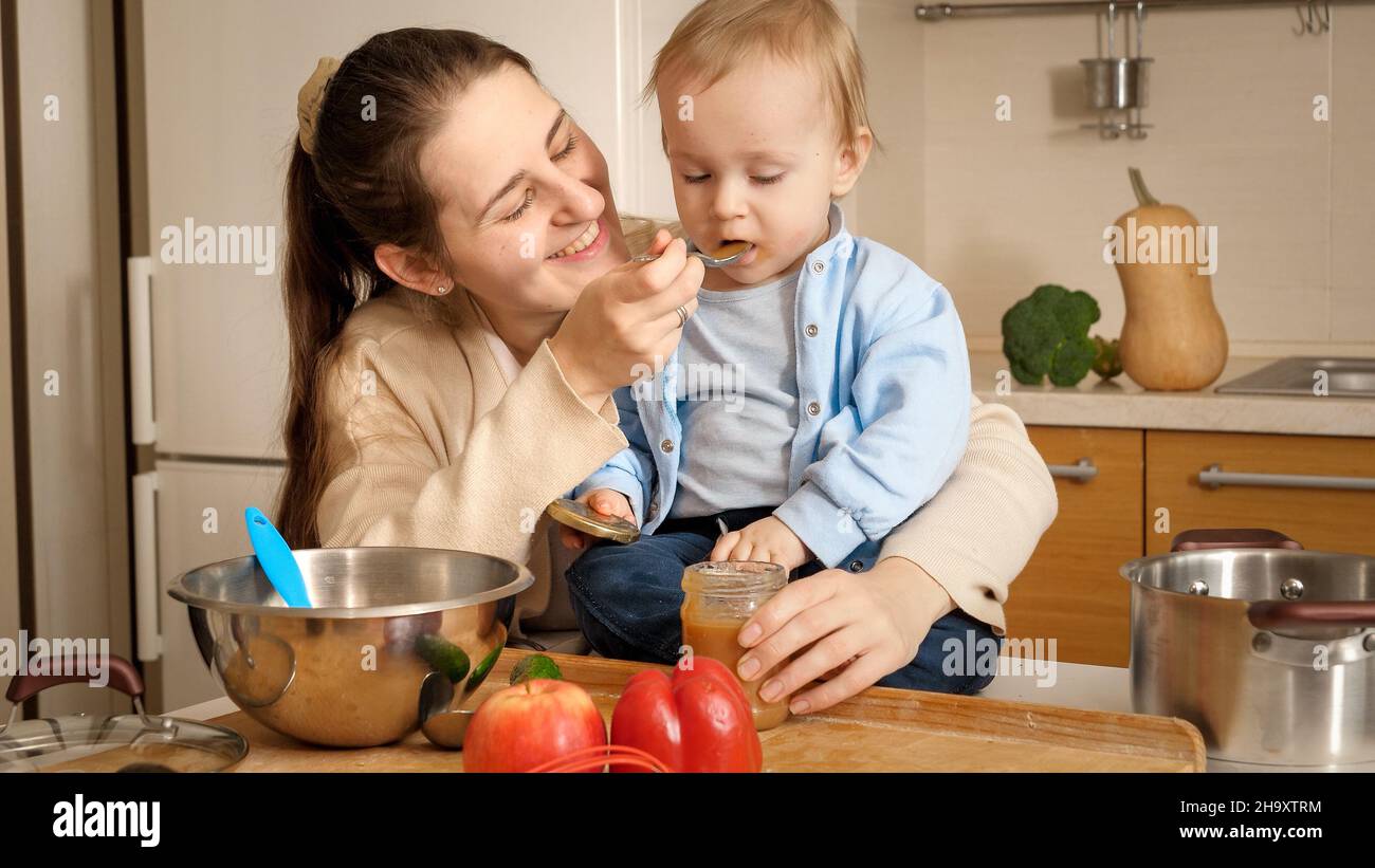 Portrait of young smiling mother feeding her baby son with fruit puree ...