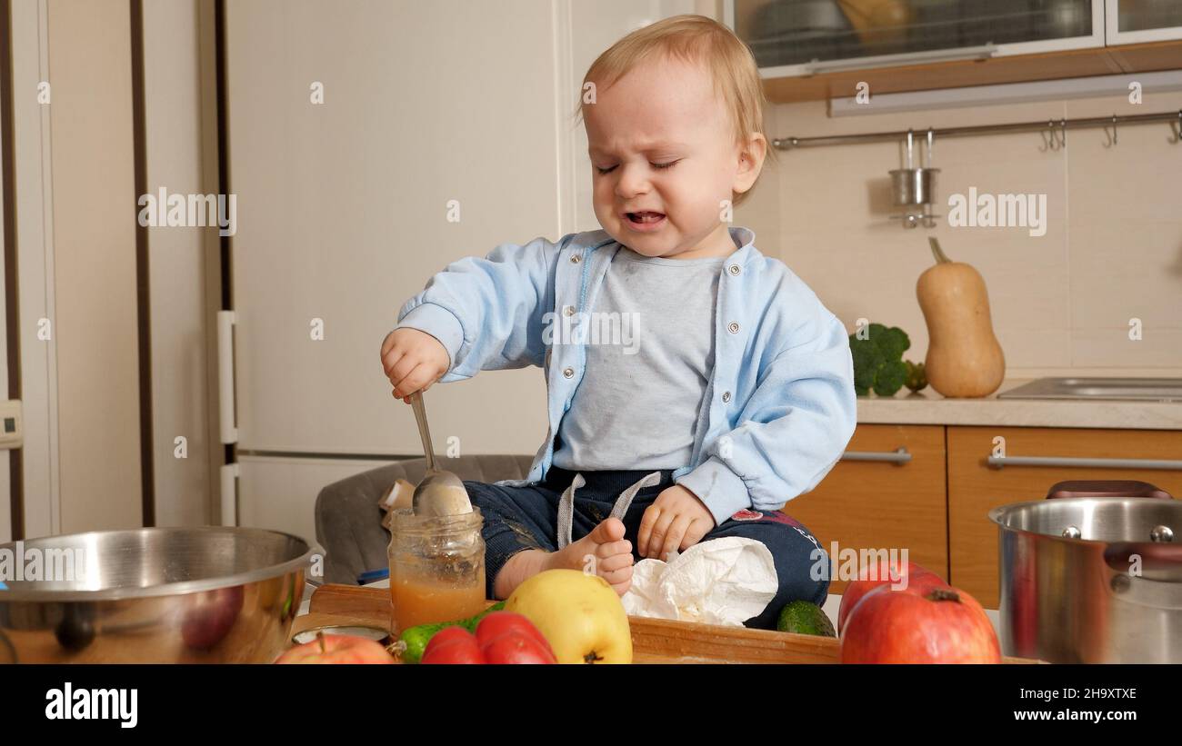 Little crying baby boy trying eating porridge with spoon on kitchen ...