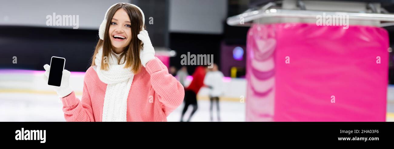 happy woman in ear muffs holding smartphone with blank screen, banner ...