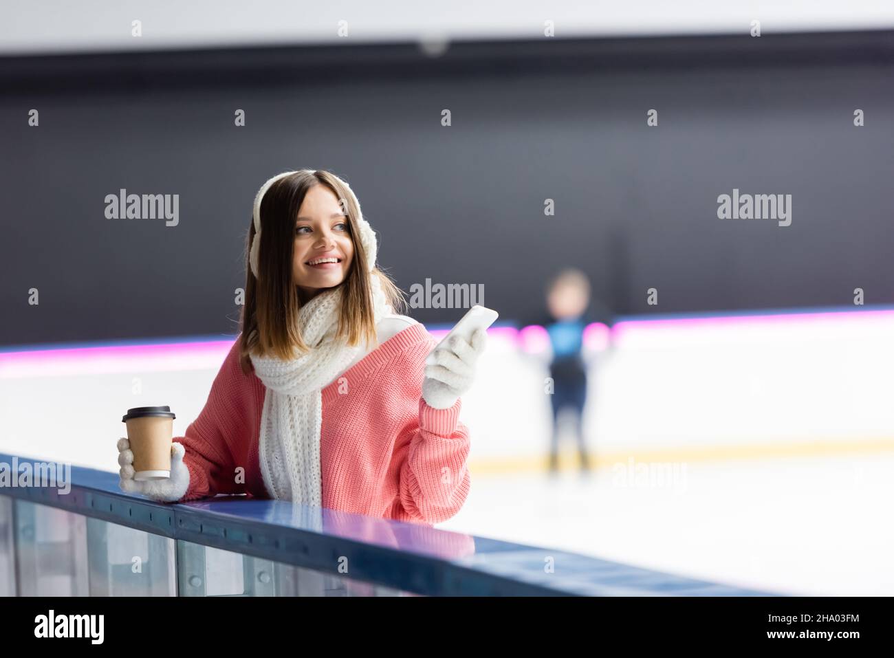 pleased woman in white ear muffs and pink sweater holding paper cup and ...
