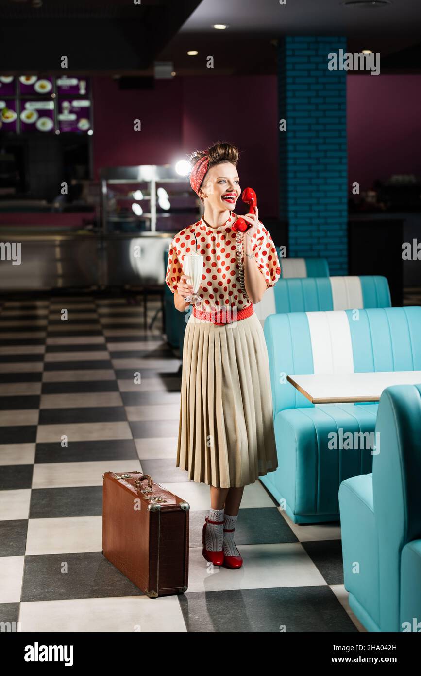 happy young pin up woman holding milkshake and handset near retro ...