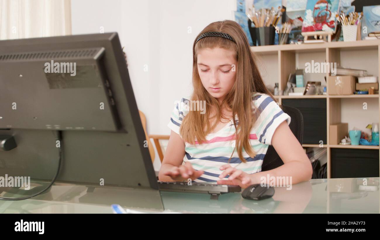 Schoolgirl sitting at desk table in front of computer typing online ...