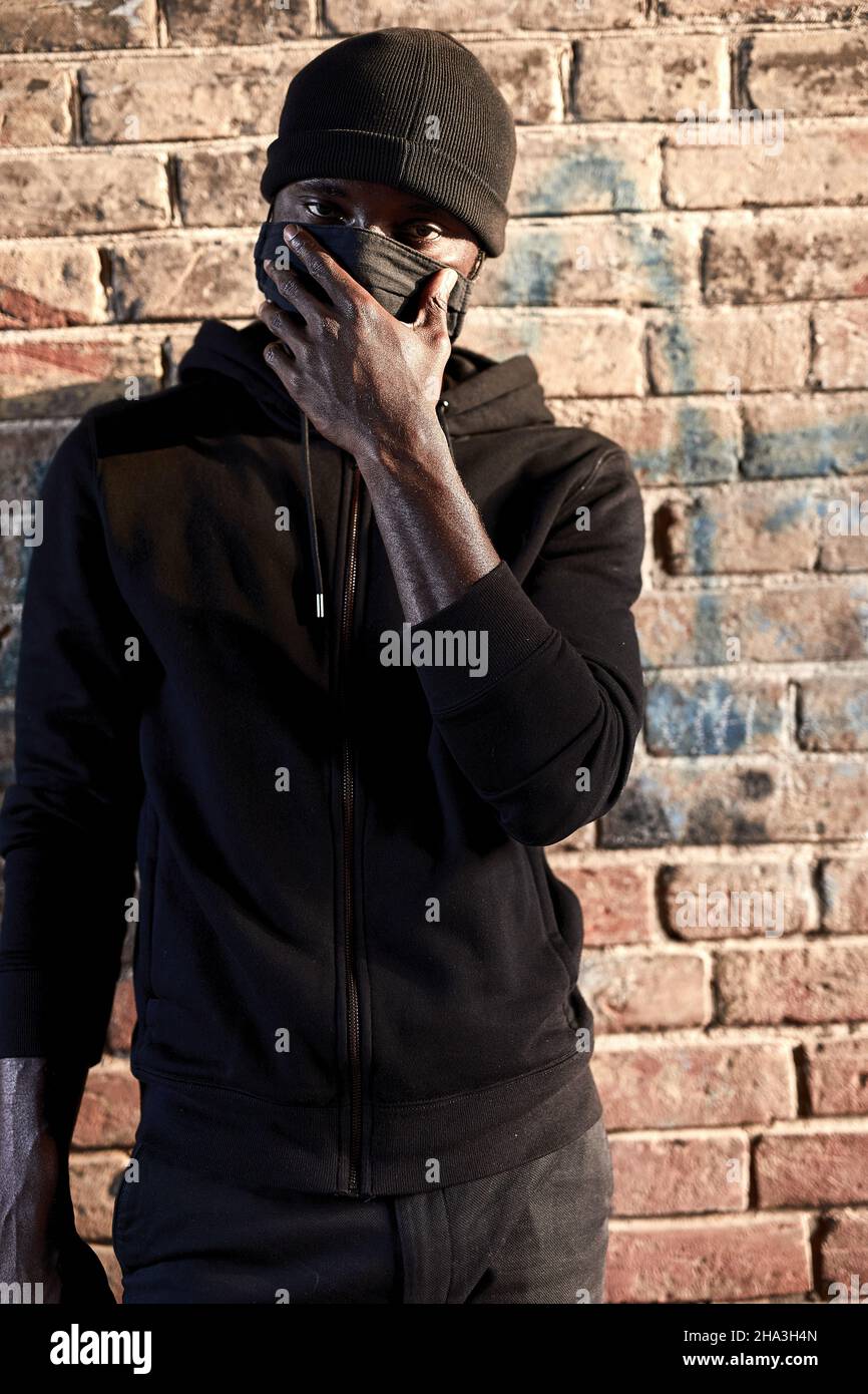 Portrait of young african man in mask standing by brick wall, hooligan ...