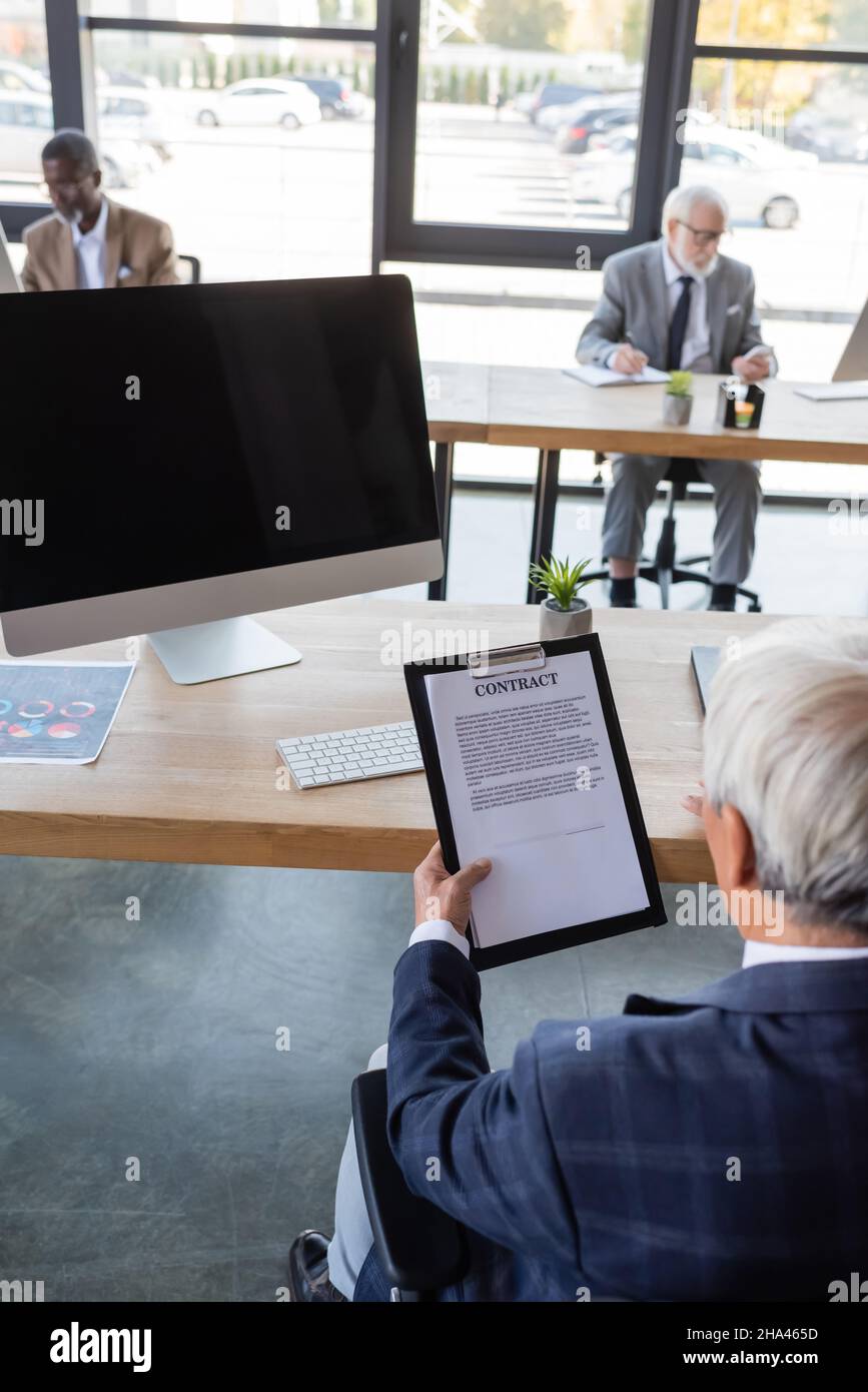 senior businessman reading contract while sitting near computer monitor ...