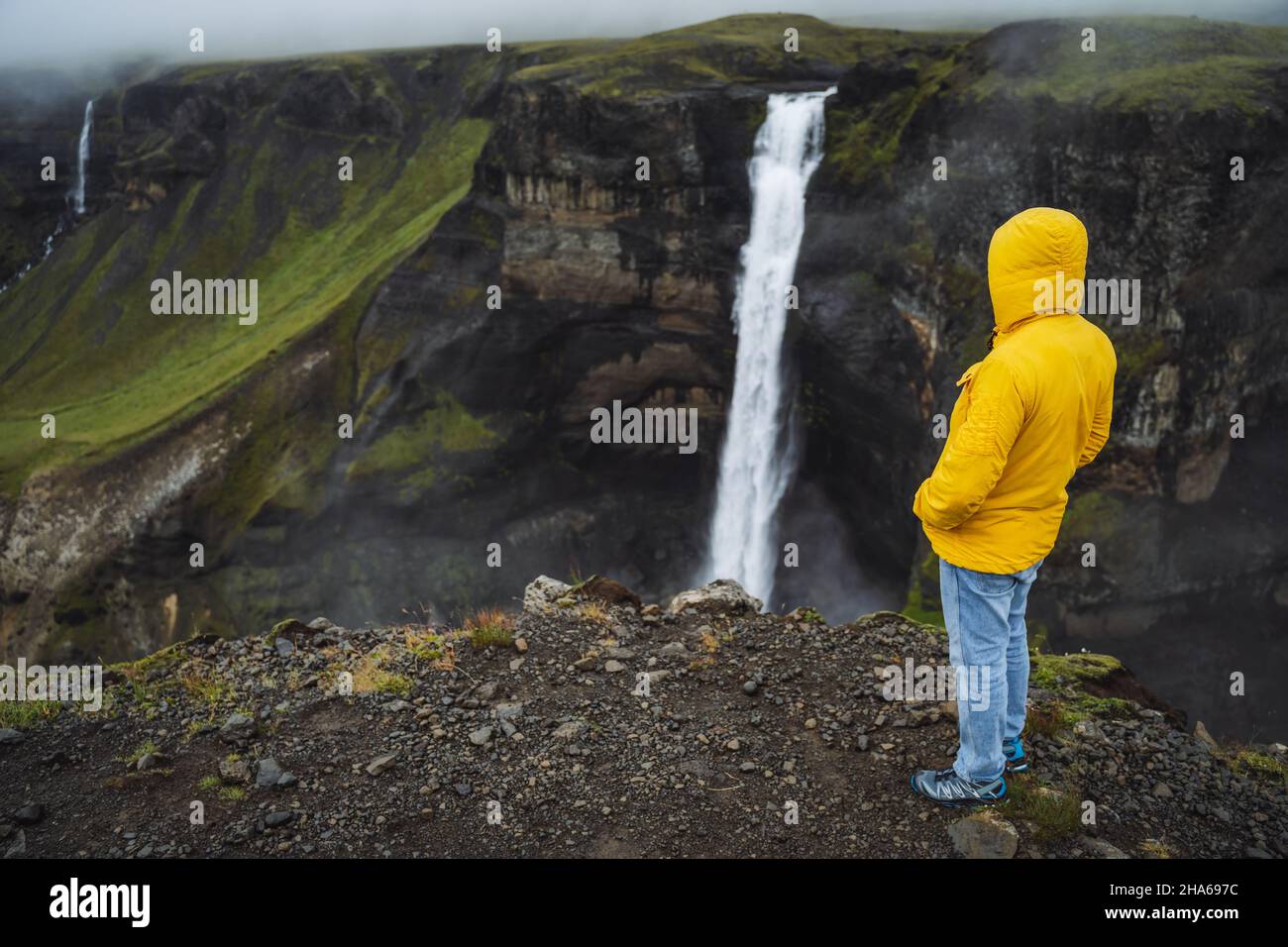 A man in yellow jacket enjoying Haifoss waterfall on rainy overcast ...