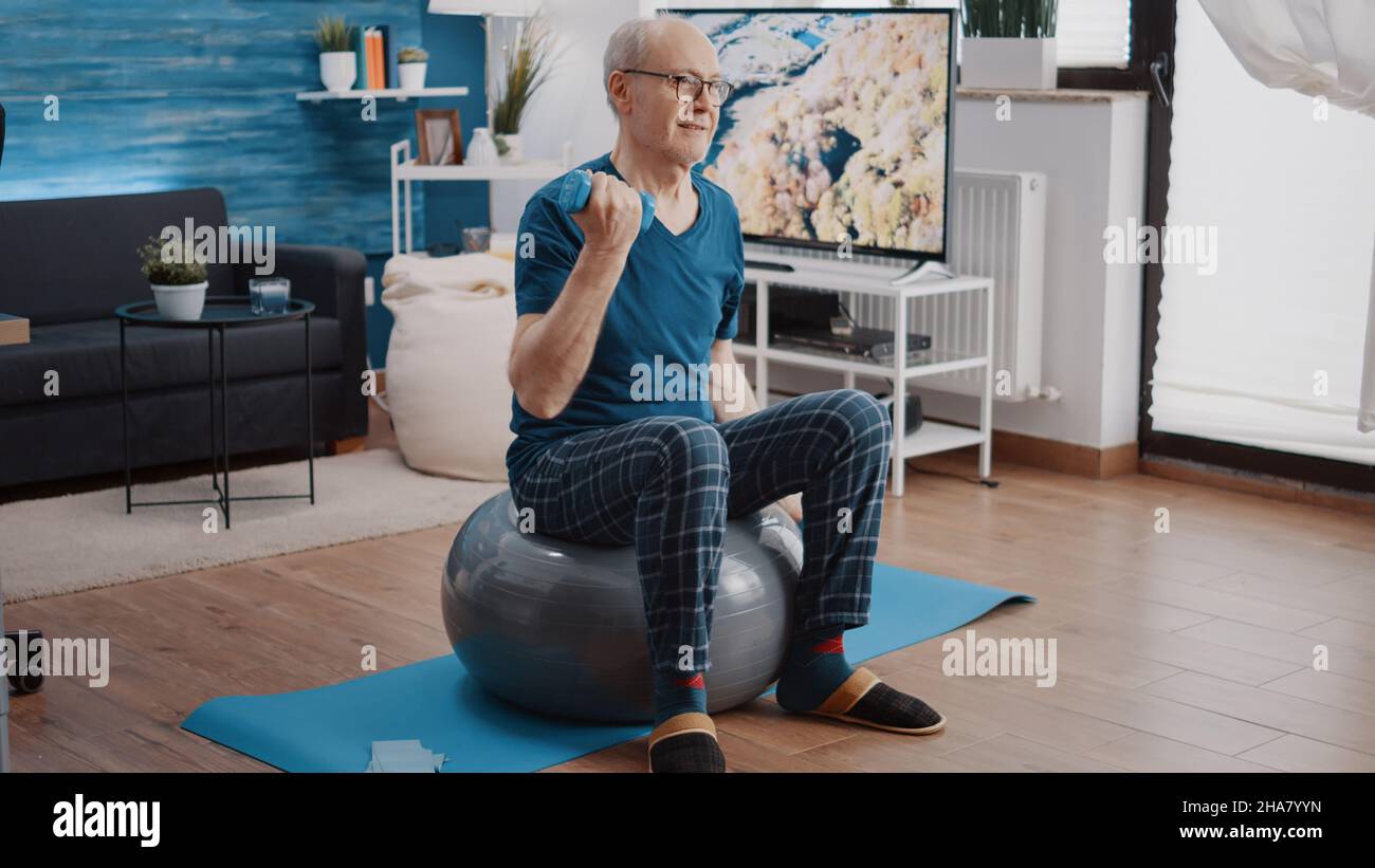 Senior man lifting dumbbells and sitting on fitness toning ball to do ...