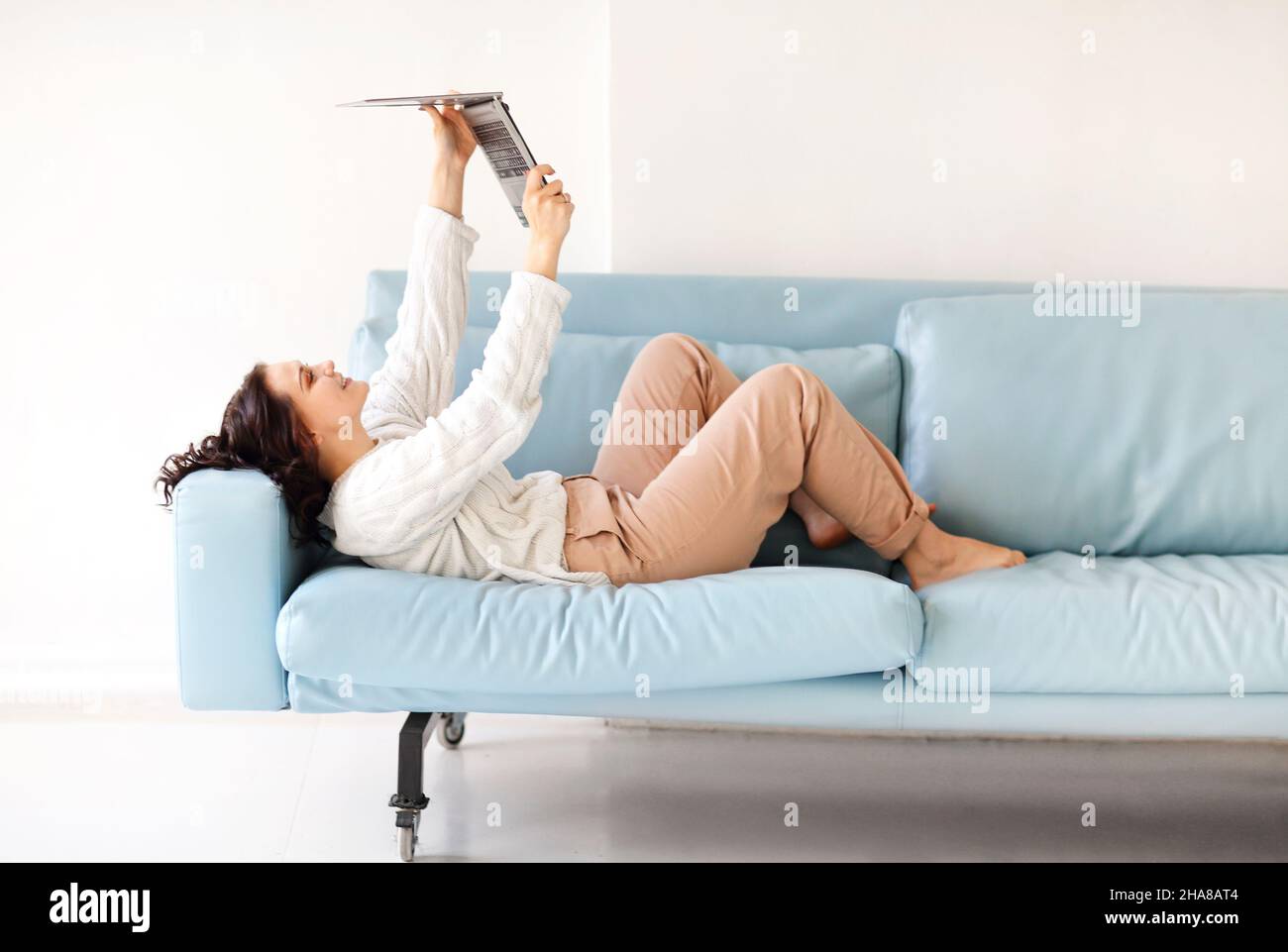 Happy young woman lying down on cozy sofa at home holding open laptop ...