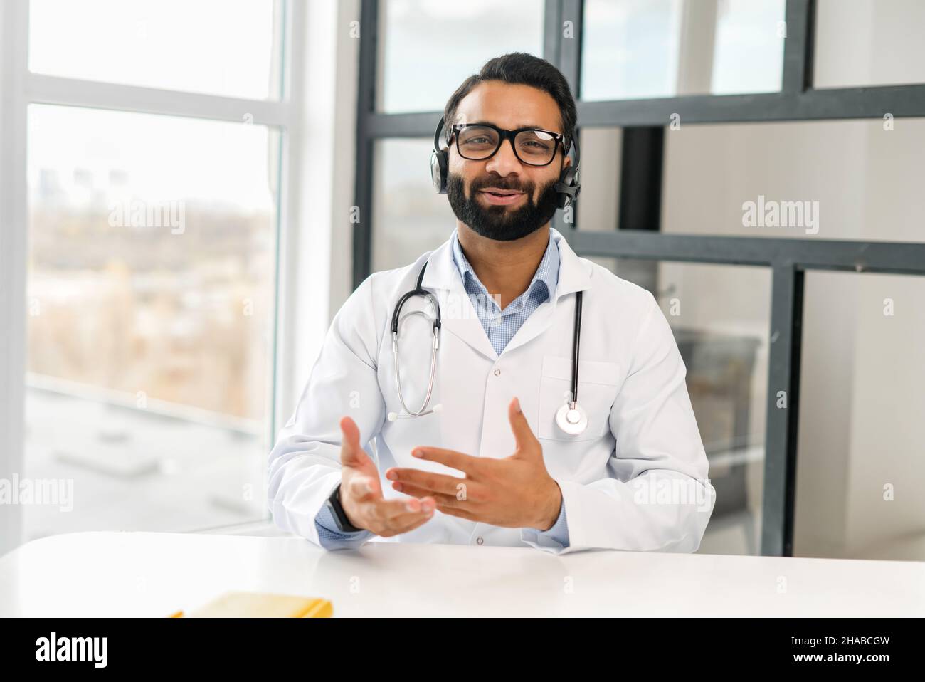 Confident Indian male MD wearing headset, eyeglasses and medical gown ...